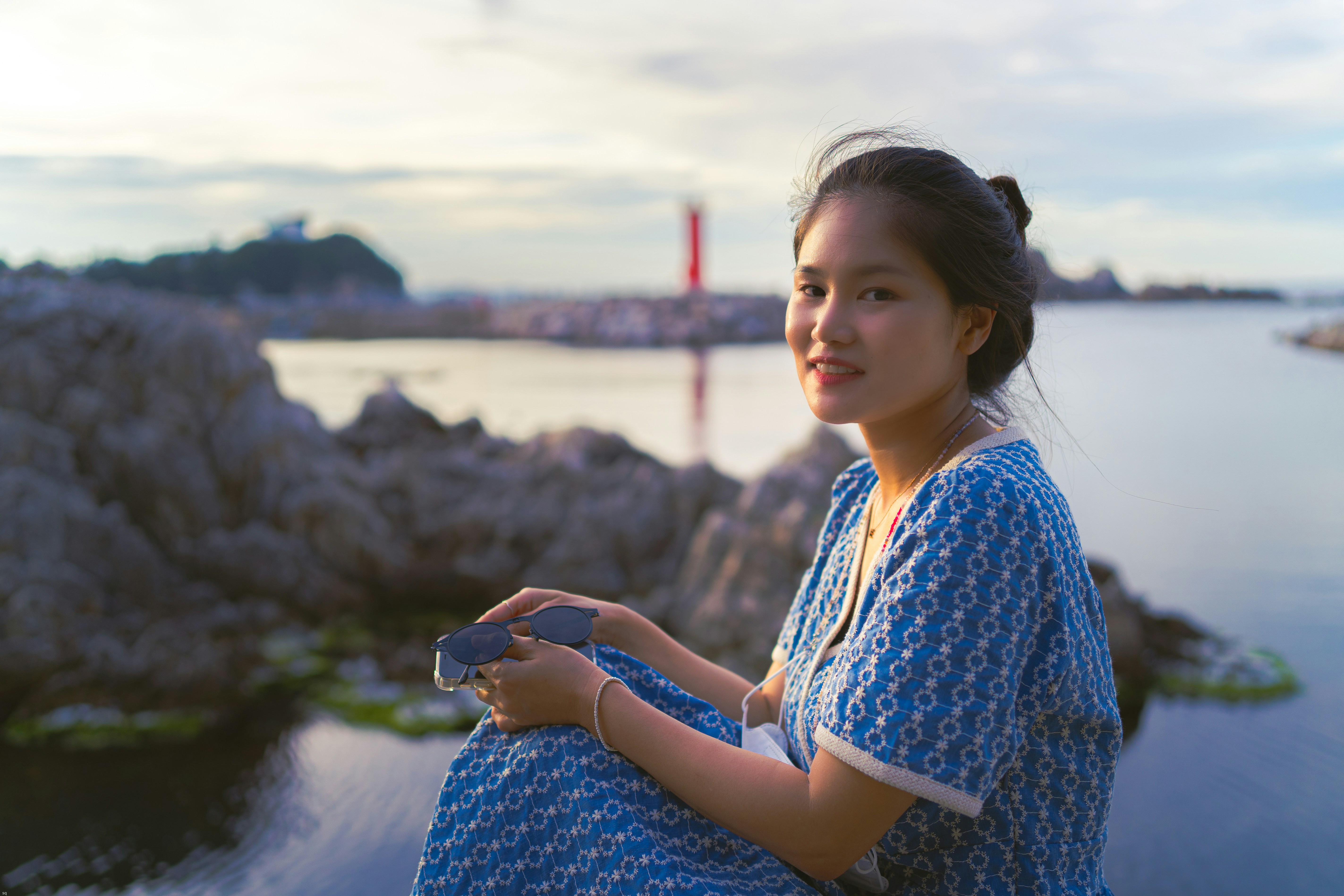 A girl in a blue dress sitting in front of a harbor with a lighthouse.
