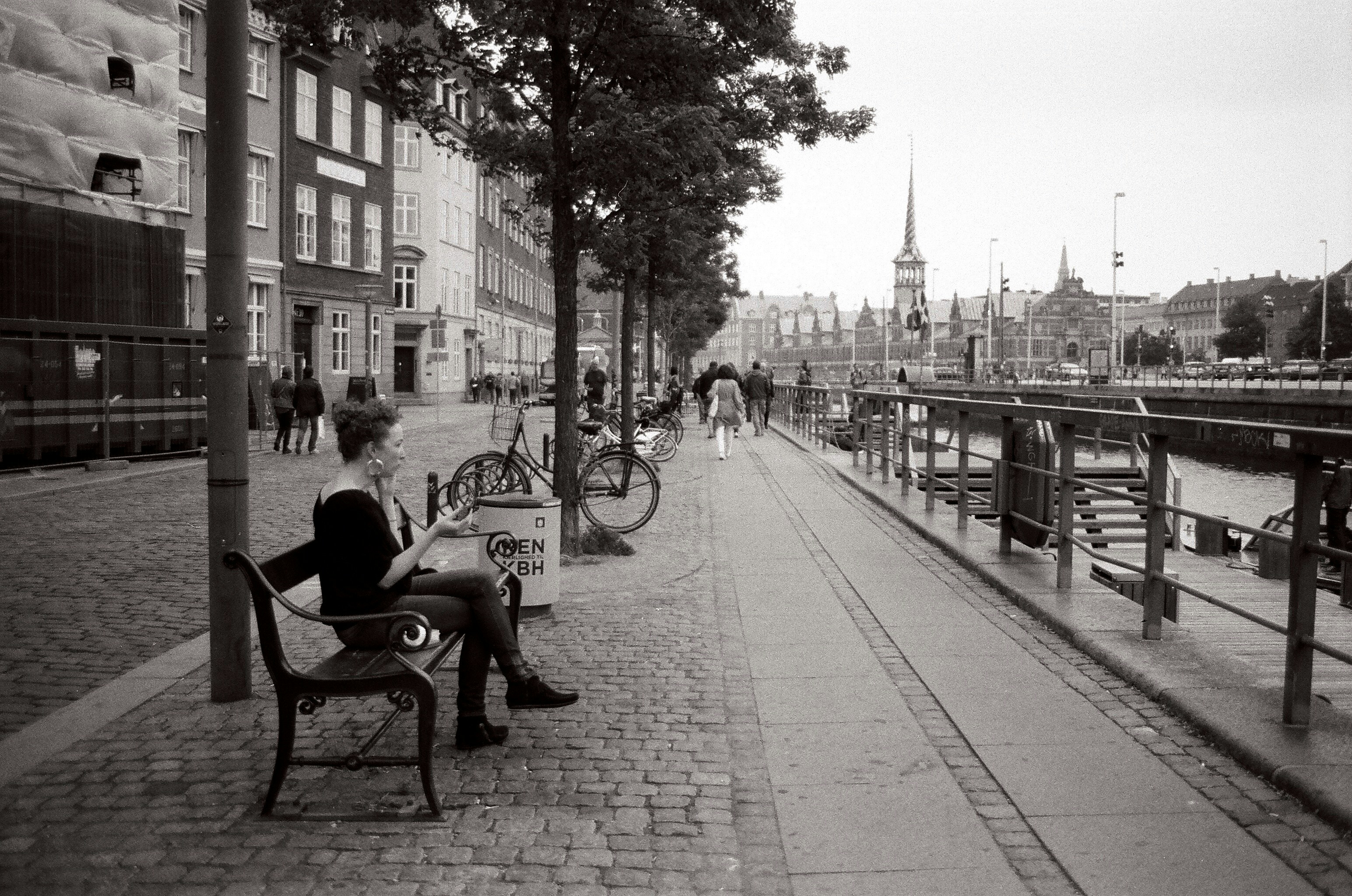 Woman sitting on a bench along a cobblestone path beside a canal, surrounded by bicycles and historical architecture. The scene evokes a sense of tranquility in an urban setting.