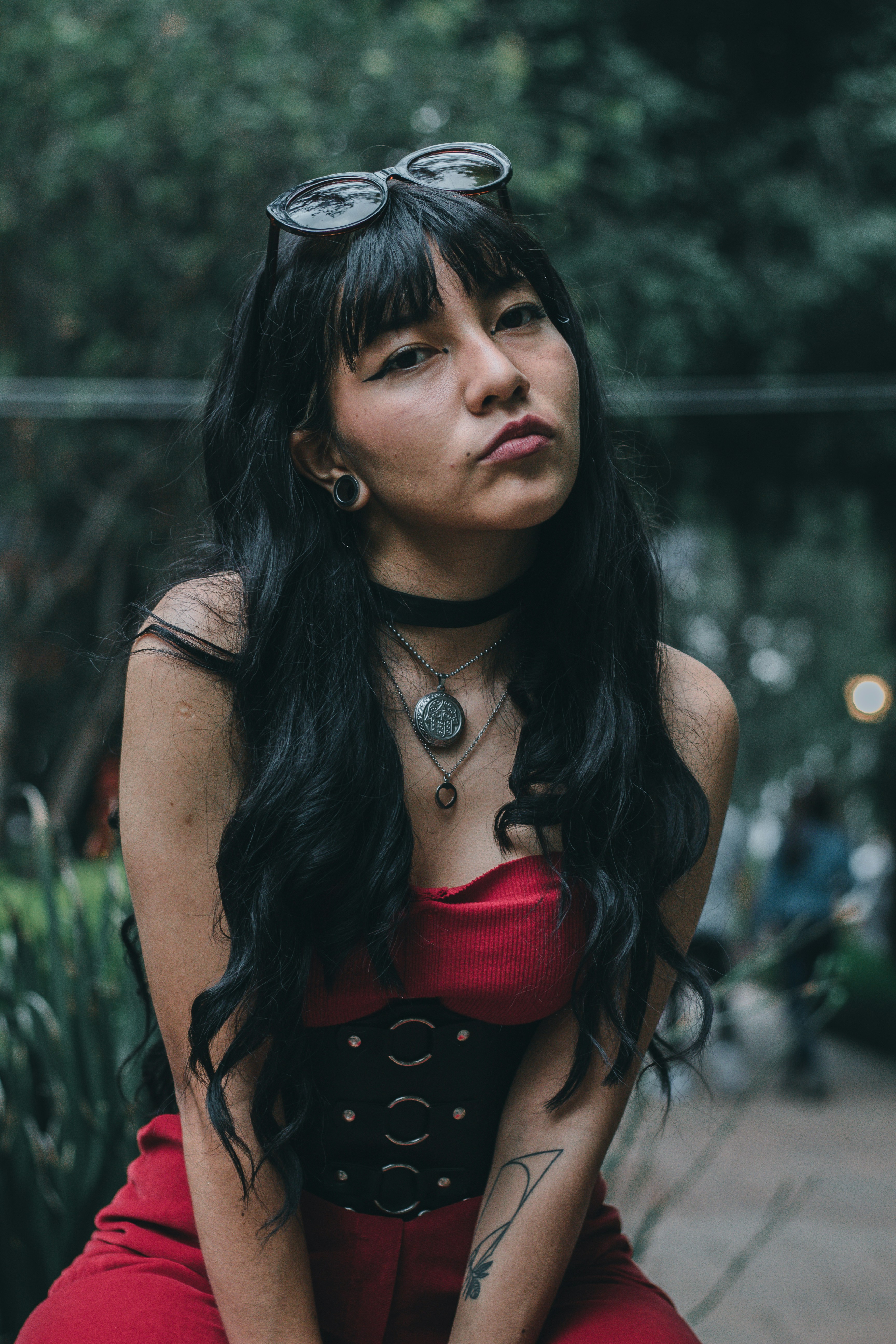 A young woman with long black hair poses confidently in a red outfit, showcasing her unique style amidst a blurred urban backdrop.