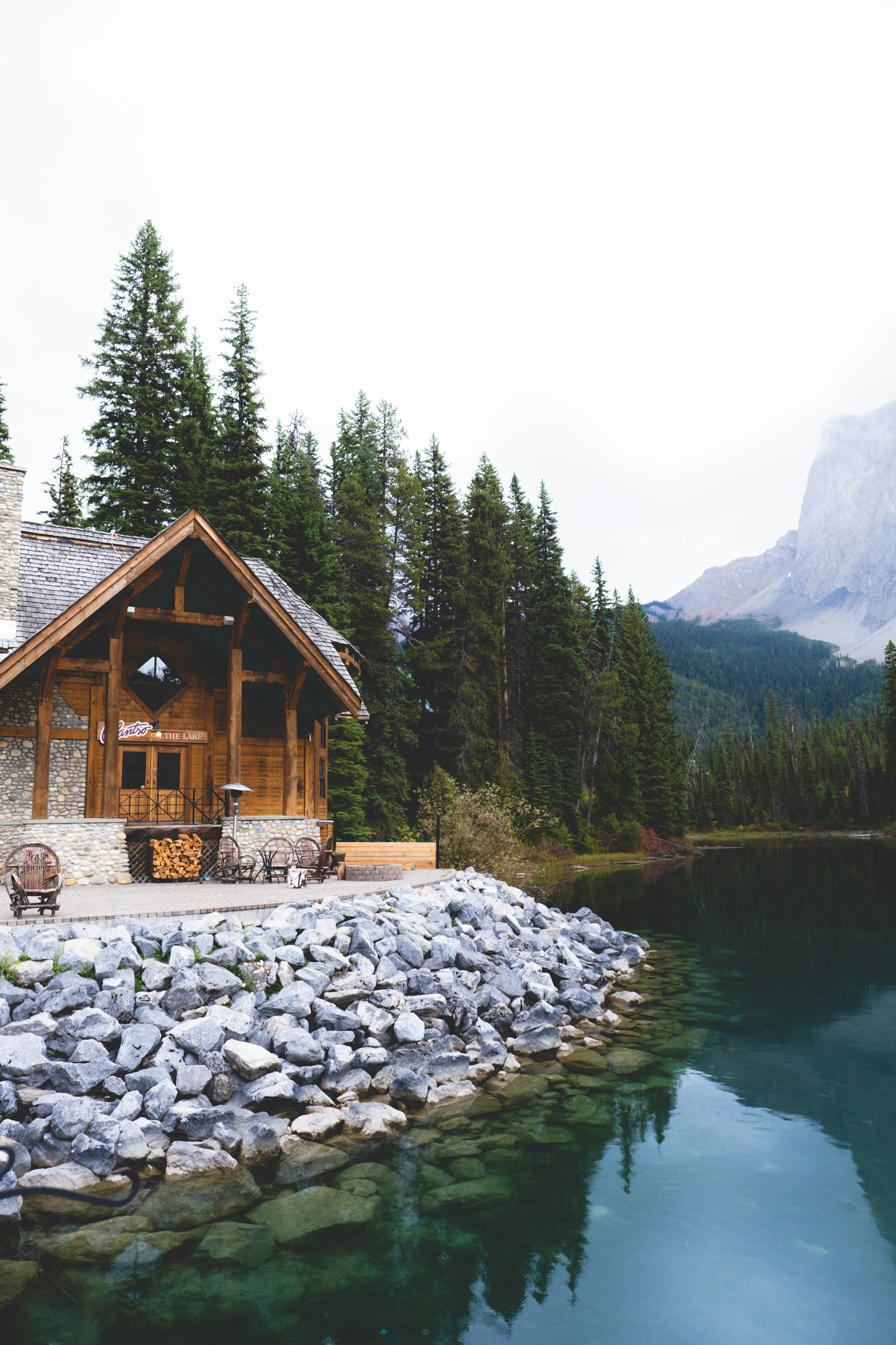 Charming wooden cabin by a tranquil lake, surrounded by lush evergreen trees and distant mountains under a cloudy sky.