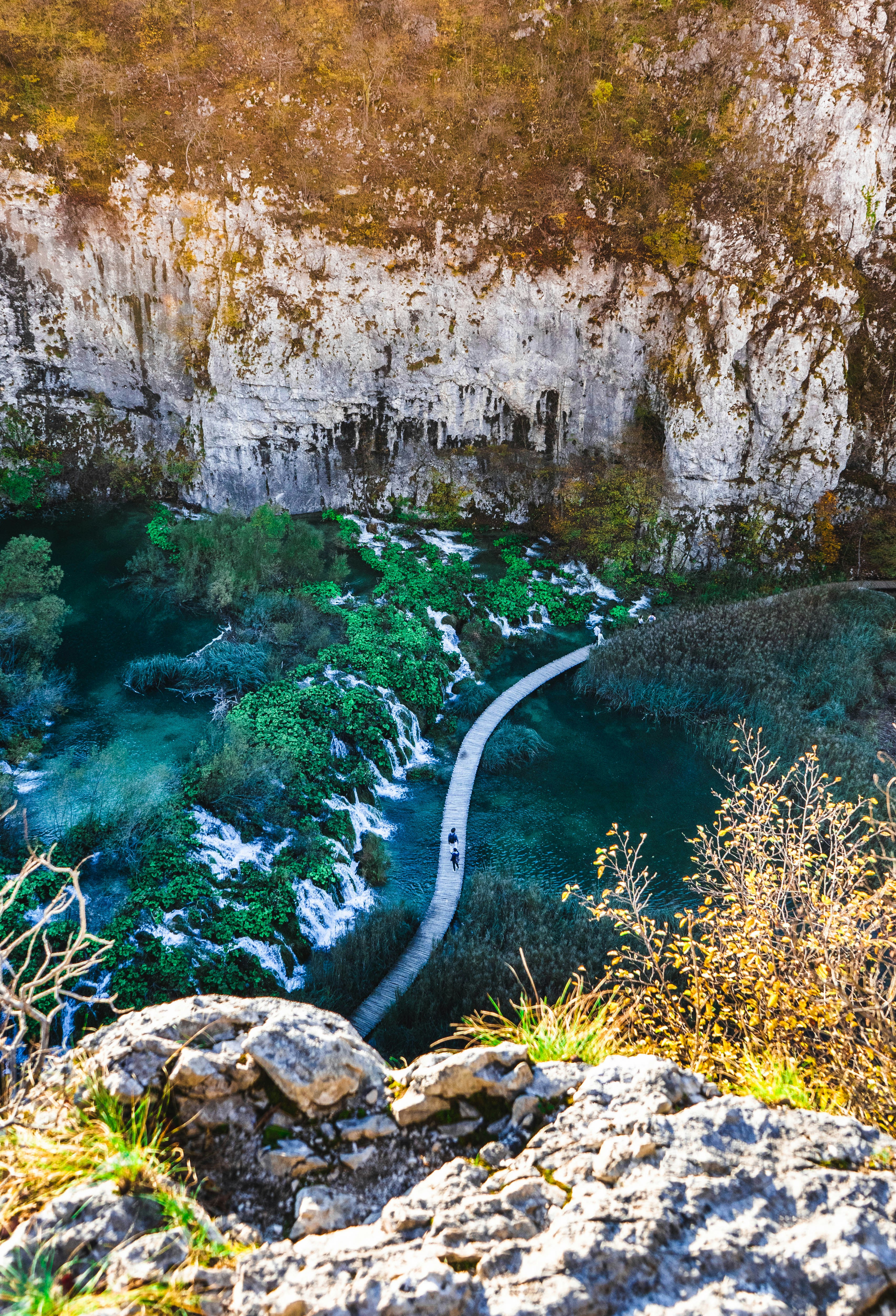A wooden walkway meanders through lush greenery and crystal-clear waters, framed by towering cliffs. The scene captures the harmony between human engineering and natural beauty.