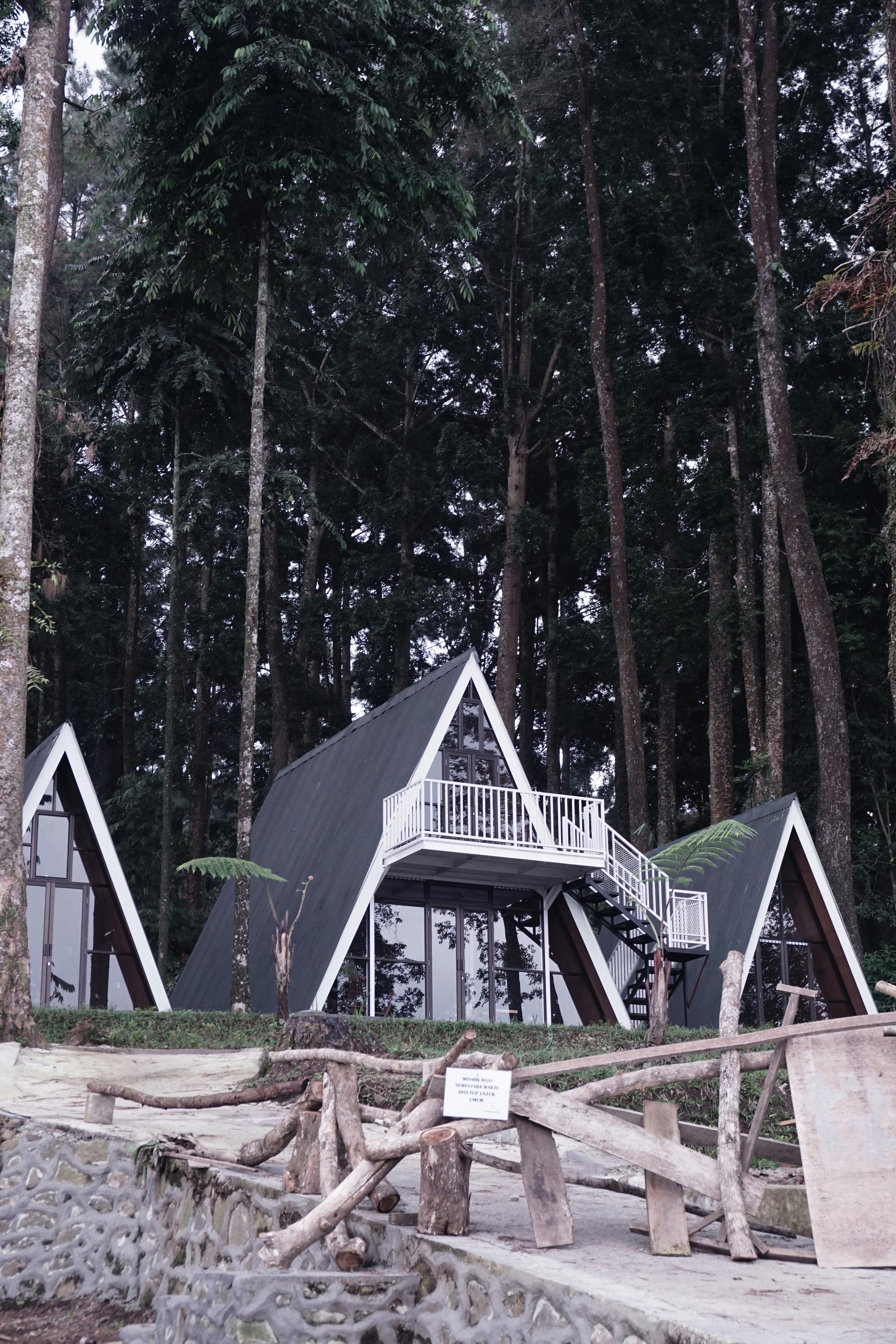 Contemporary A-frame cabin nestled among towering trees, featuring large windows and a balcony. The structure blends harmoniously with its forest surroundings.