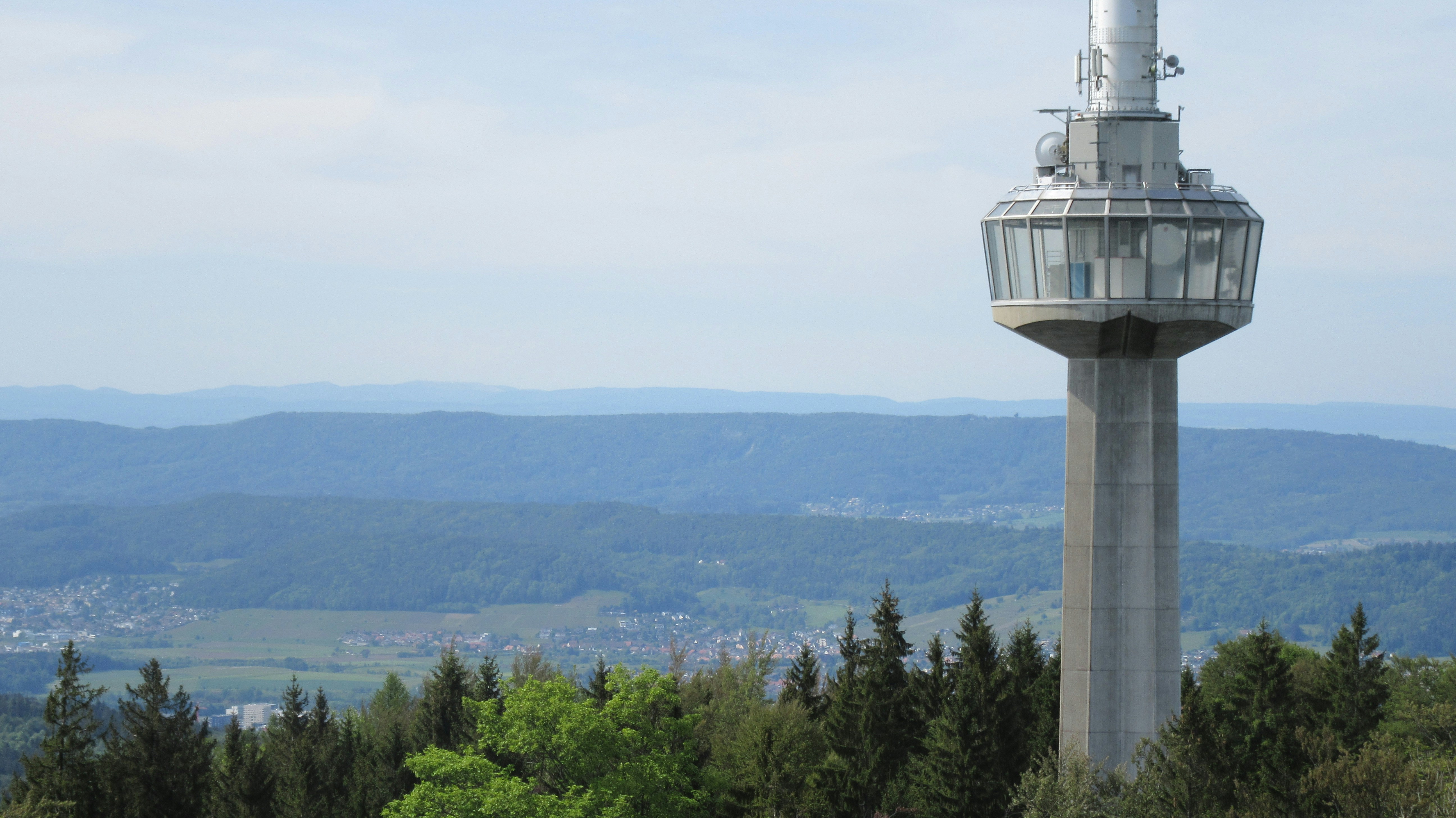 Landscape photograph featuring a tall observation tower rising on the right, framed by dense pines and distant hills. The composition emphasizes vertical contrast against a calm blue valley.