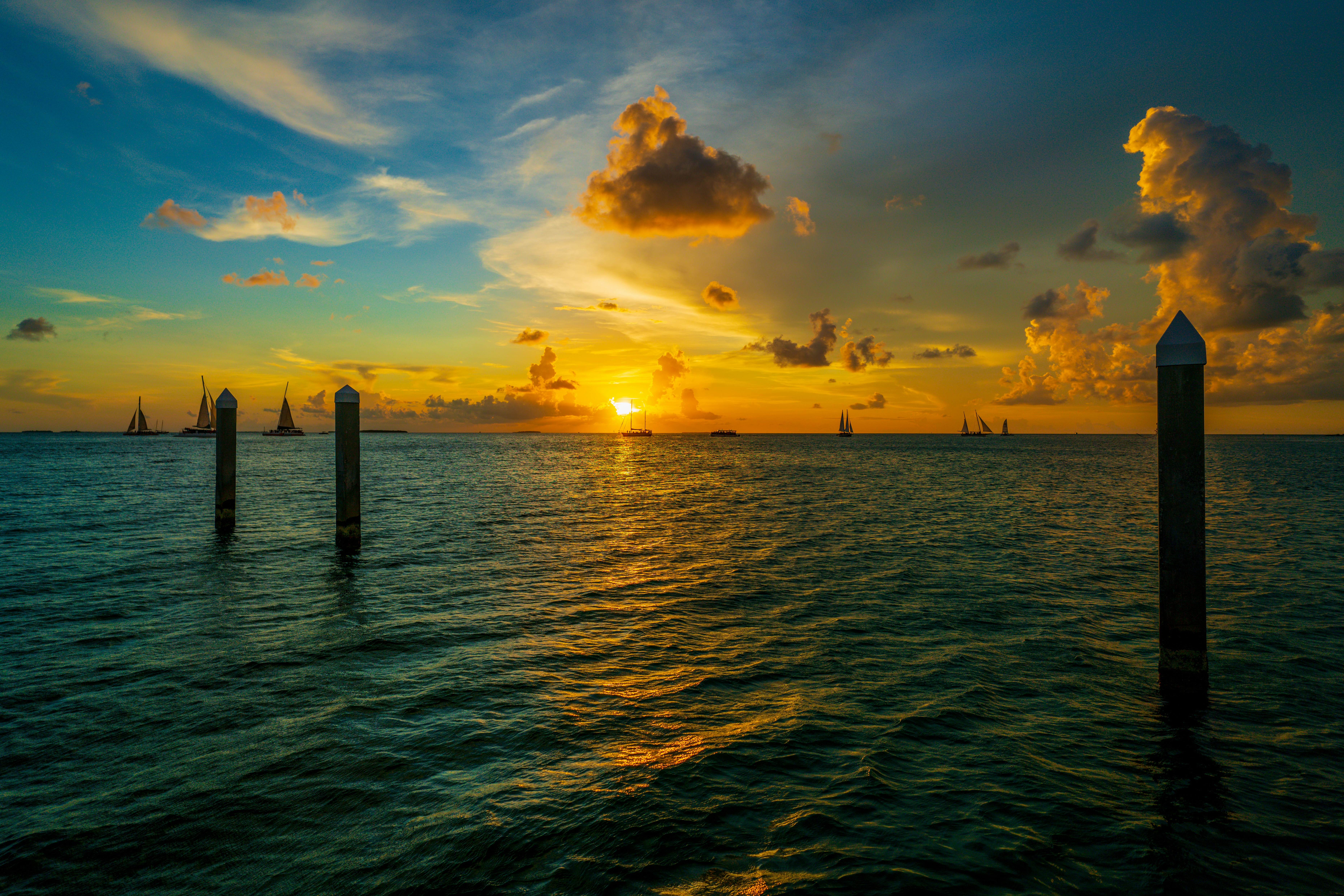 Golden sun dipping below the horizon, casting vibrant hues across the water, with sailboats gently silhouetted in the distance.