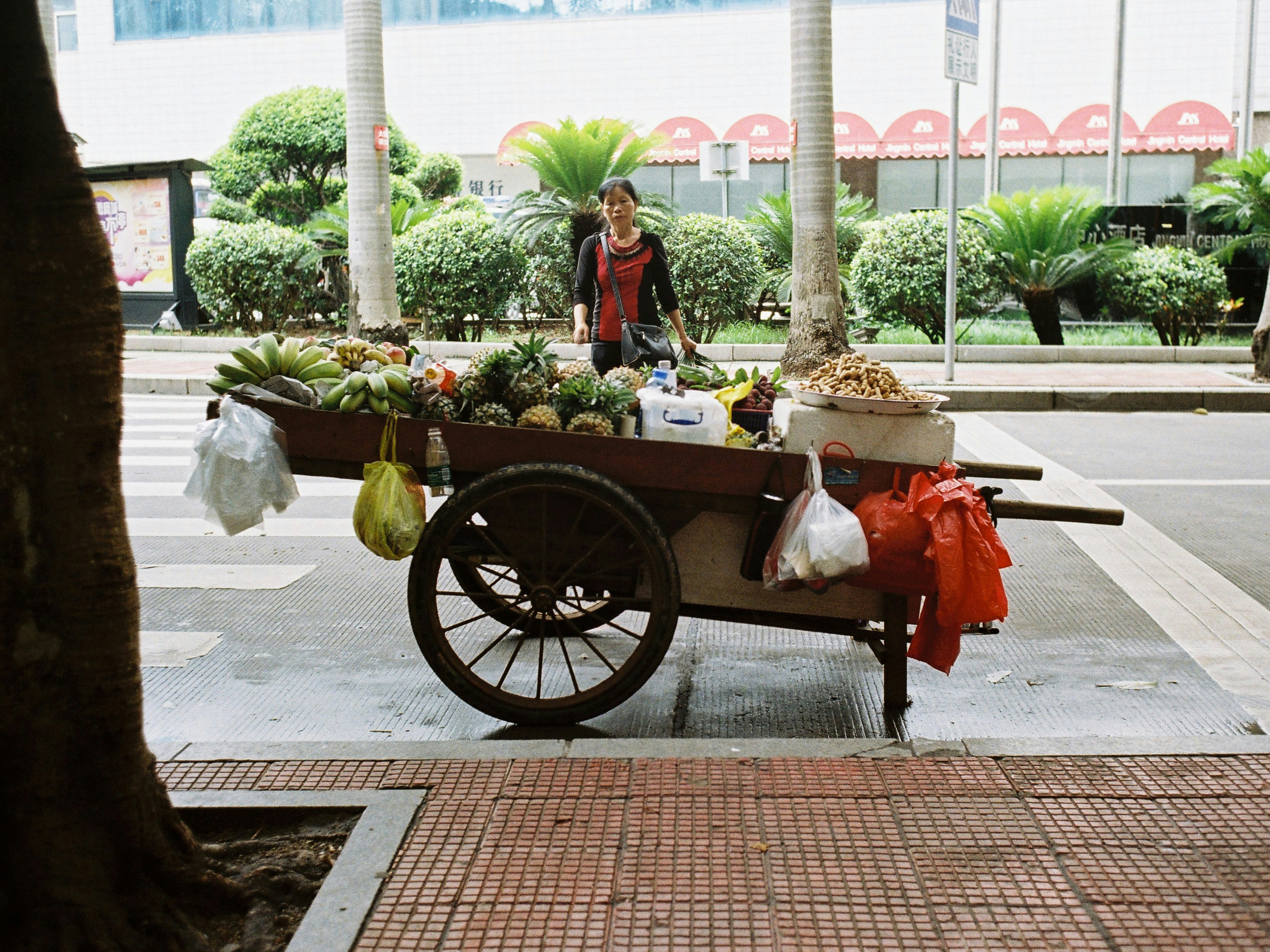 A vibrant fruit cart brimming with colorful produce sits on a bustling street, while a vendor engages with passersby. The scene encapsulates daily life in an urban setting.