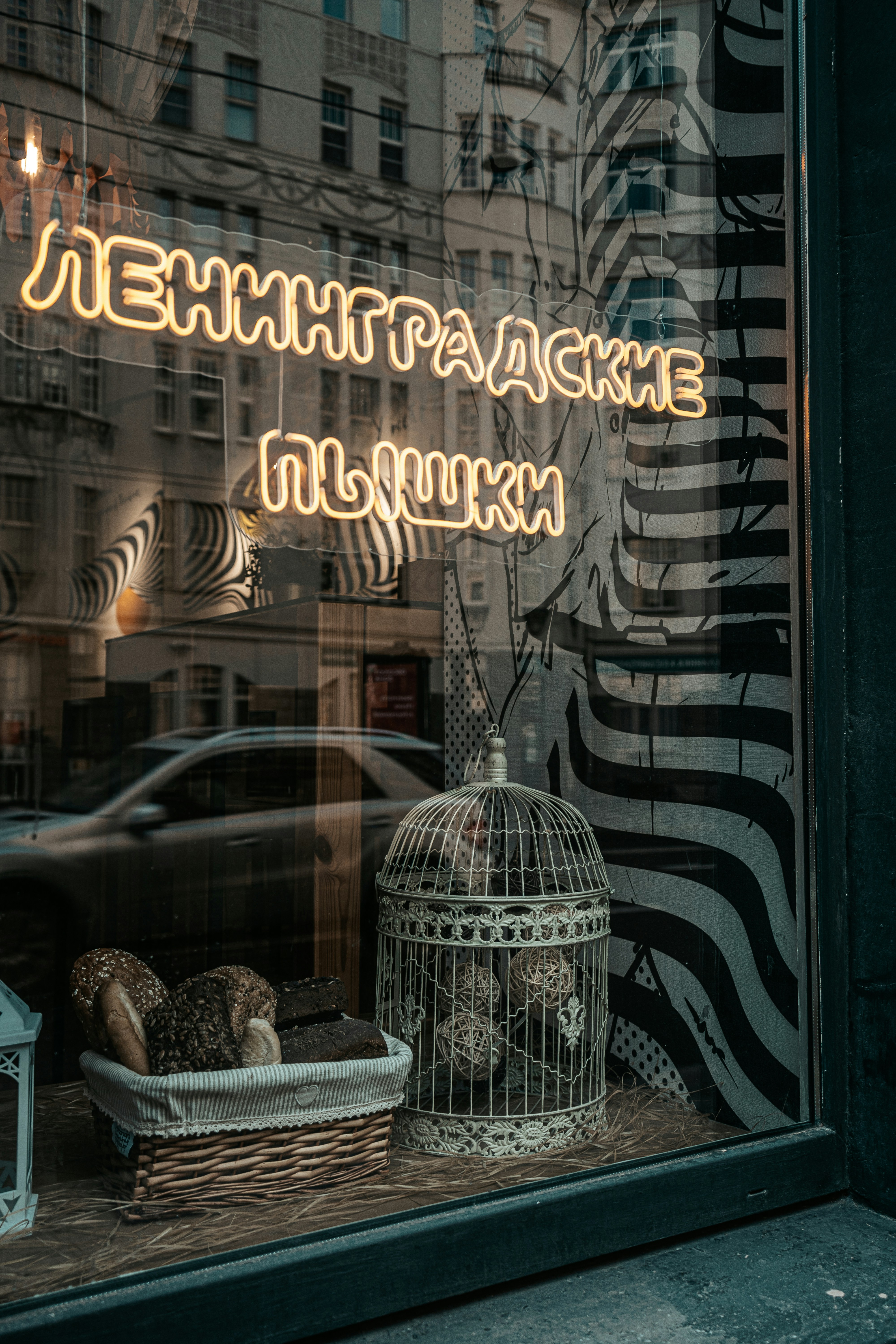 Neon Cyrillic glow over a city storefront: a white birdcage and bread basket in a reflective still life