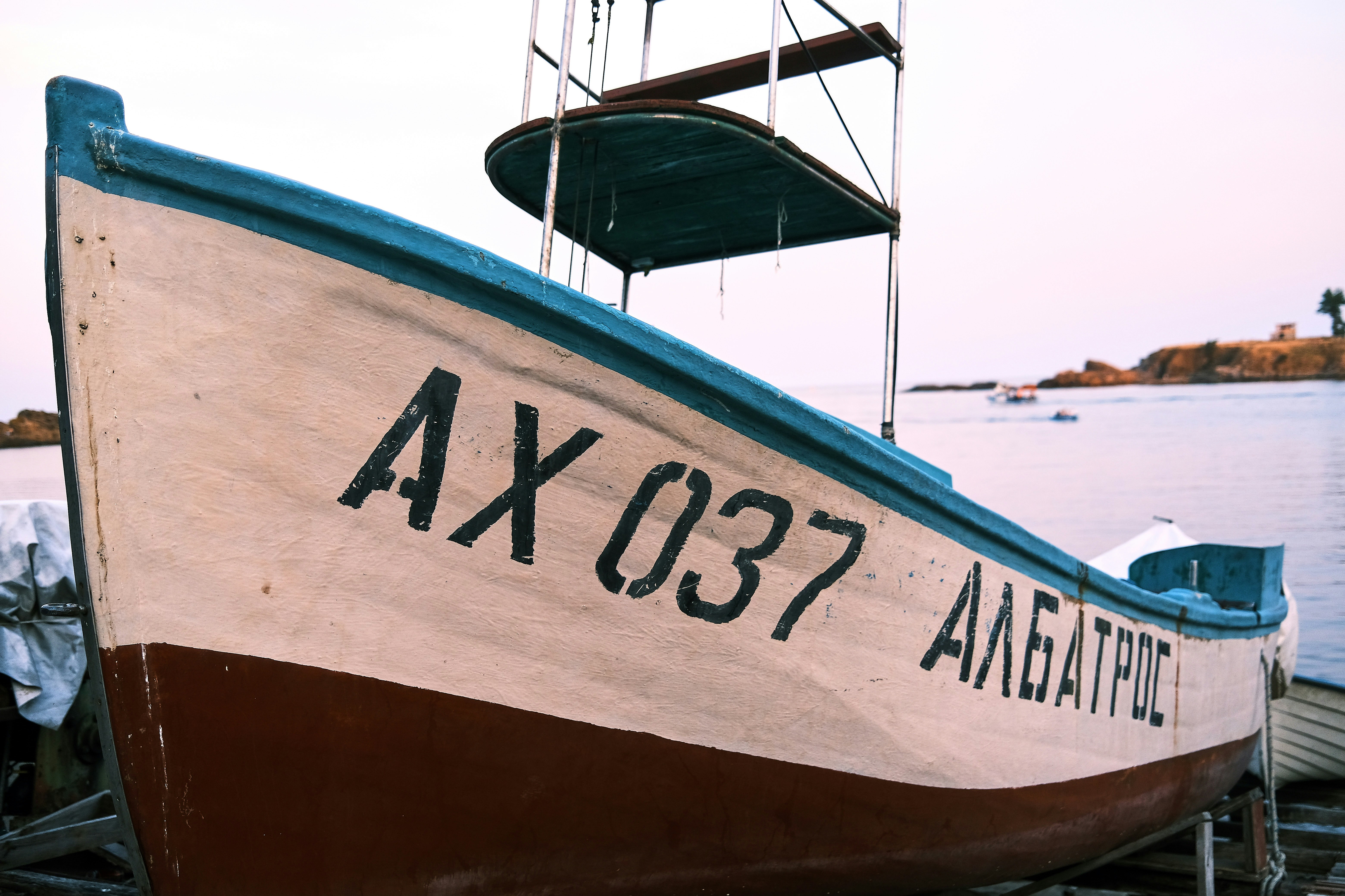 Close-up of a fishing boat with bold lettering and a rustic finish, anchored by the shore. The tranquil water reflects the soft hues of twilight.
