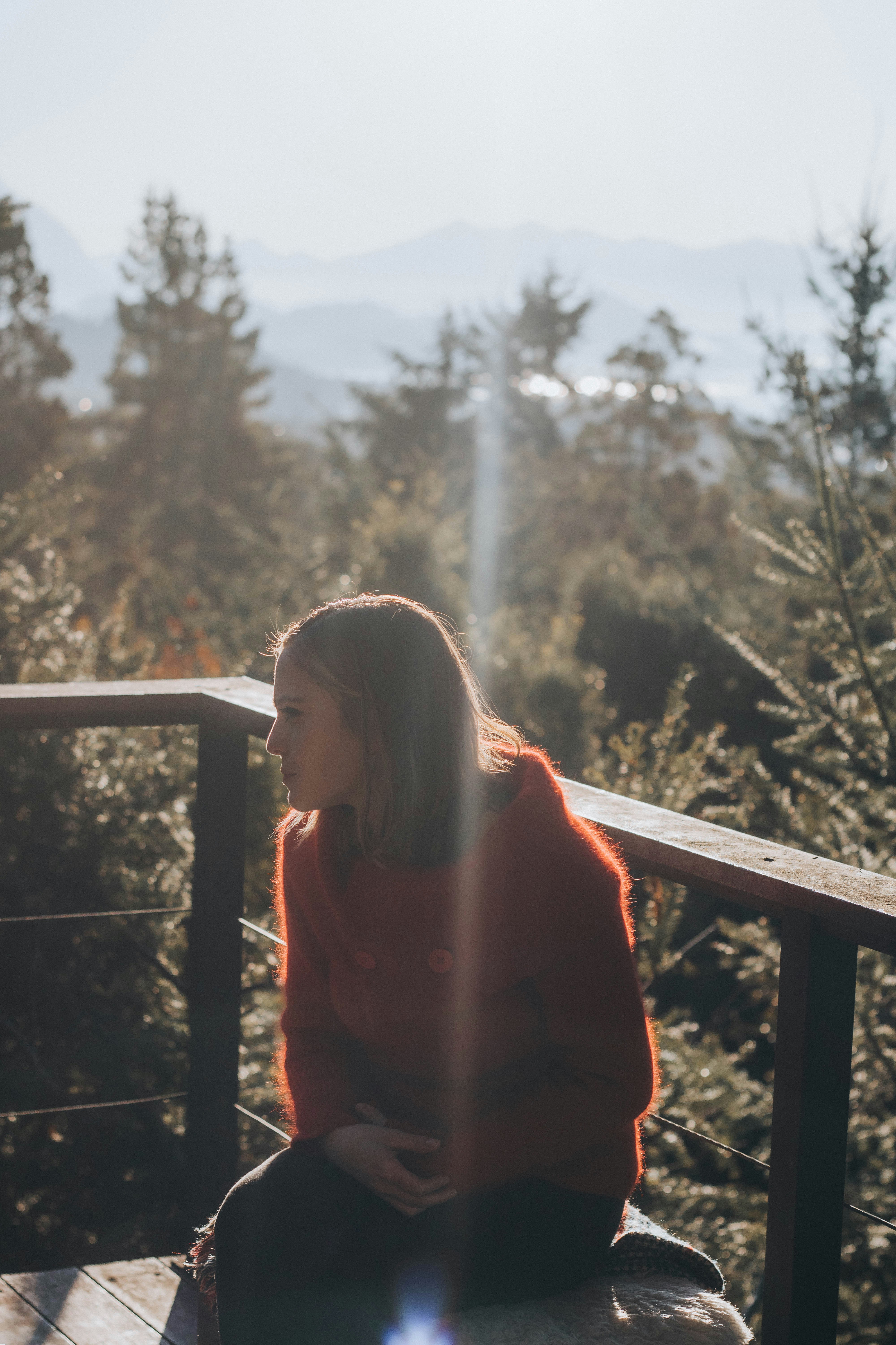 A young woman in a red sweater sits pensively on a balcony, surrounded by lush greenery and distant mountains, bathed in soft sunlight.