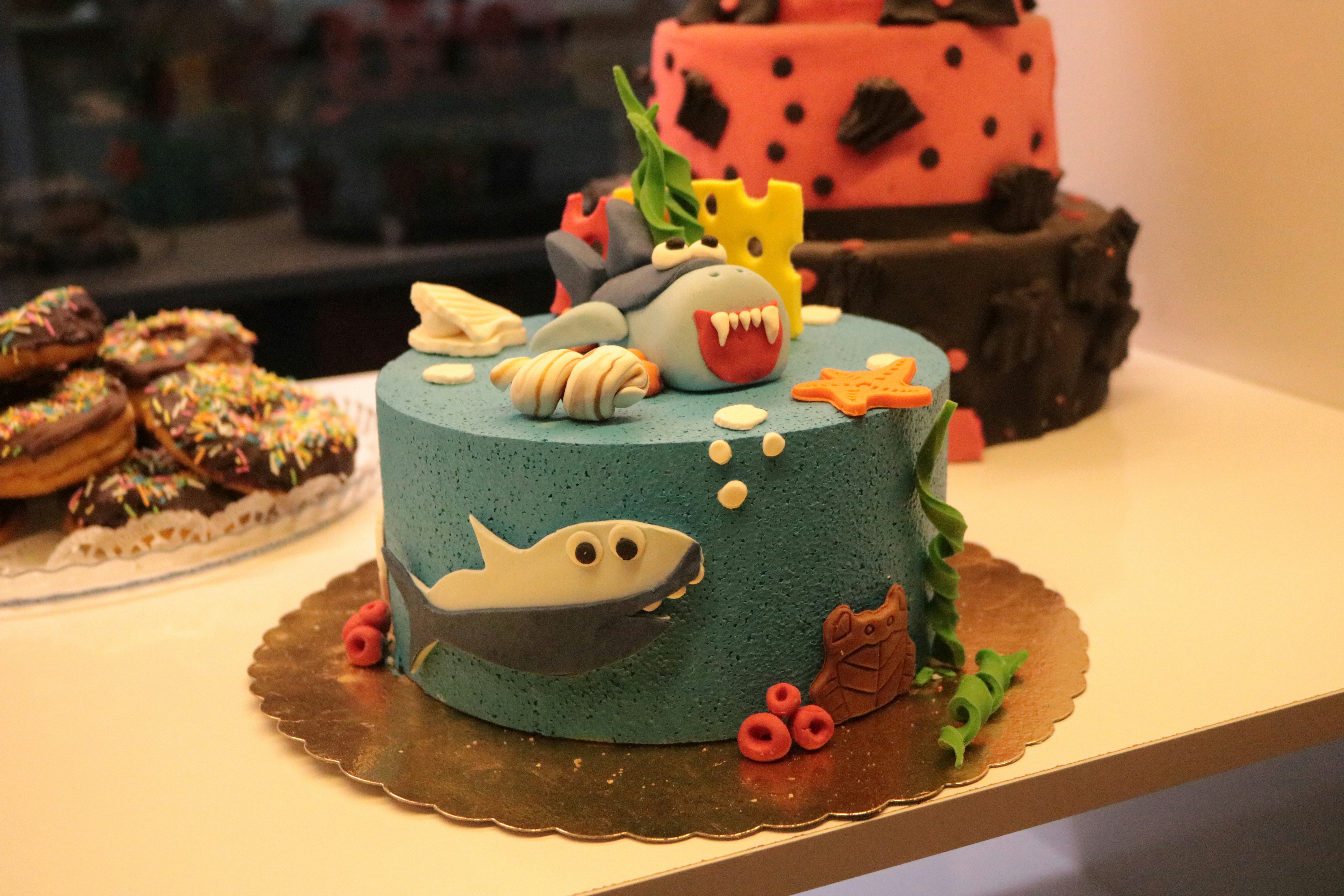 A vibrant cake decorated with playful ocean-themed elements, including a shark, starfish, and seaweed, set against a backdrop of assorted donuts.