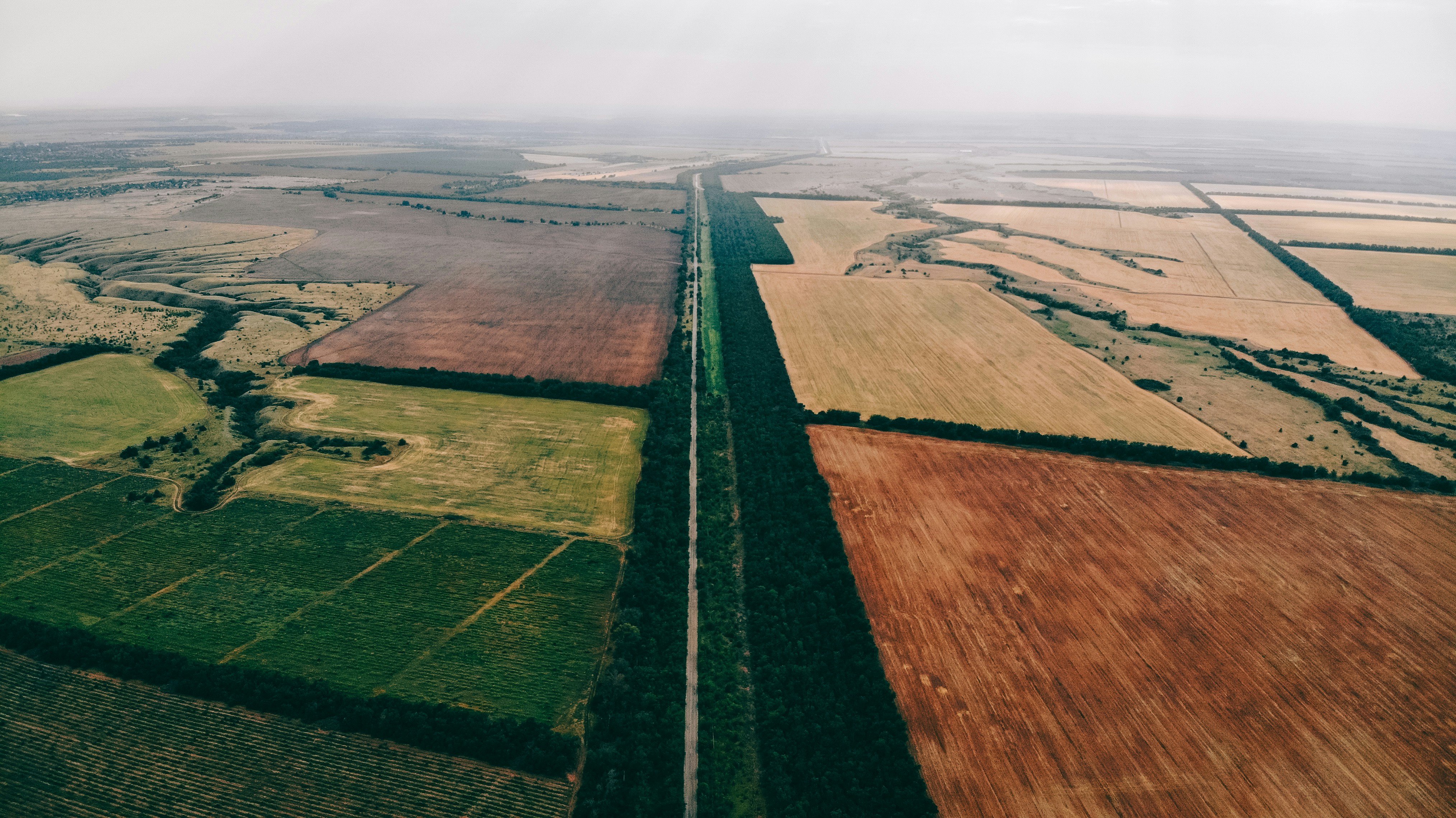 Expansive patchwork of green and brown fields under a cloudy sky, intersected by a straight road.