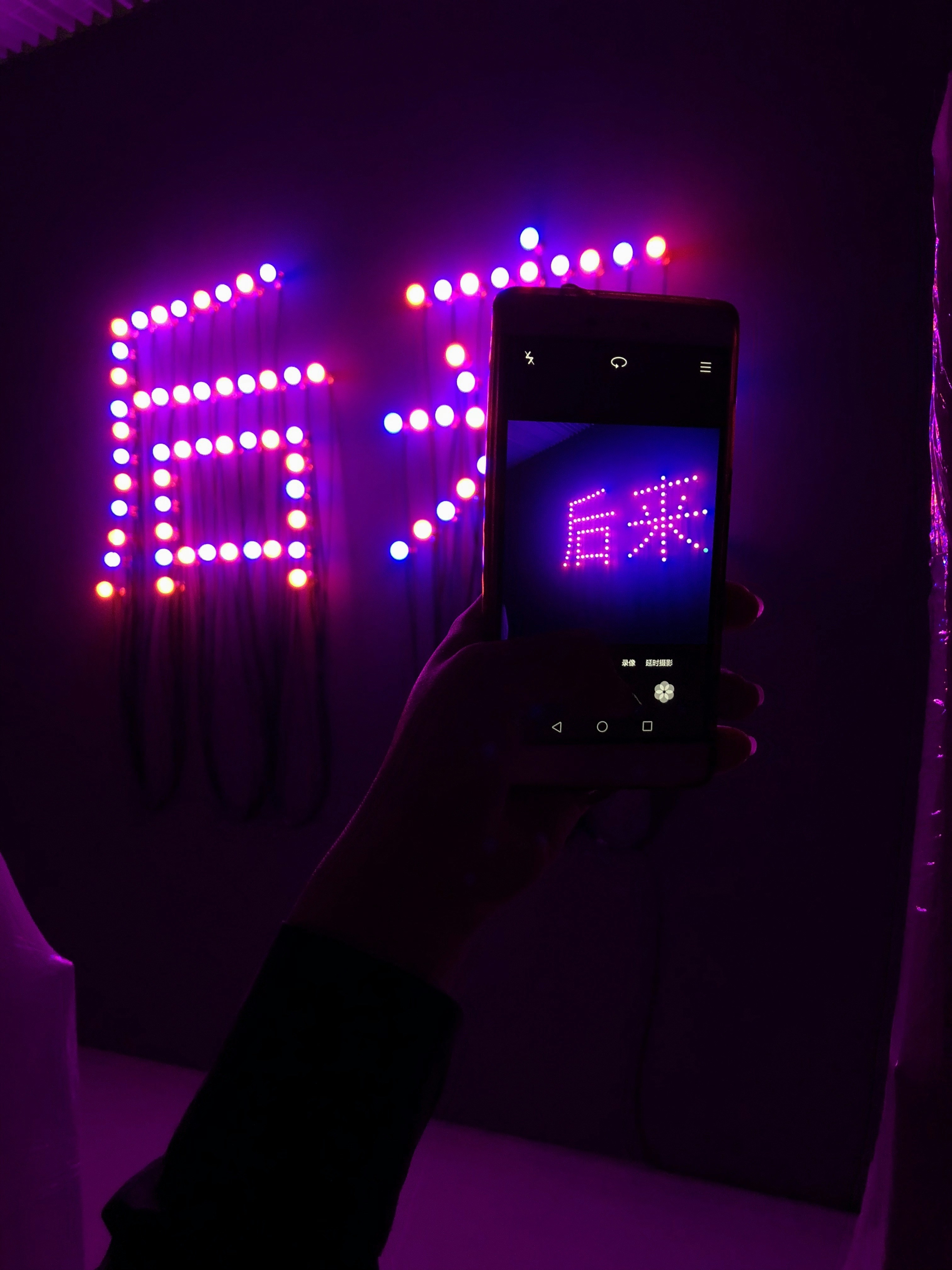 Dim room lit by purple neon; a hand holds a smartphone displaying glowing kanji, the screen catching the sign's vibrant LEDs.