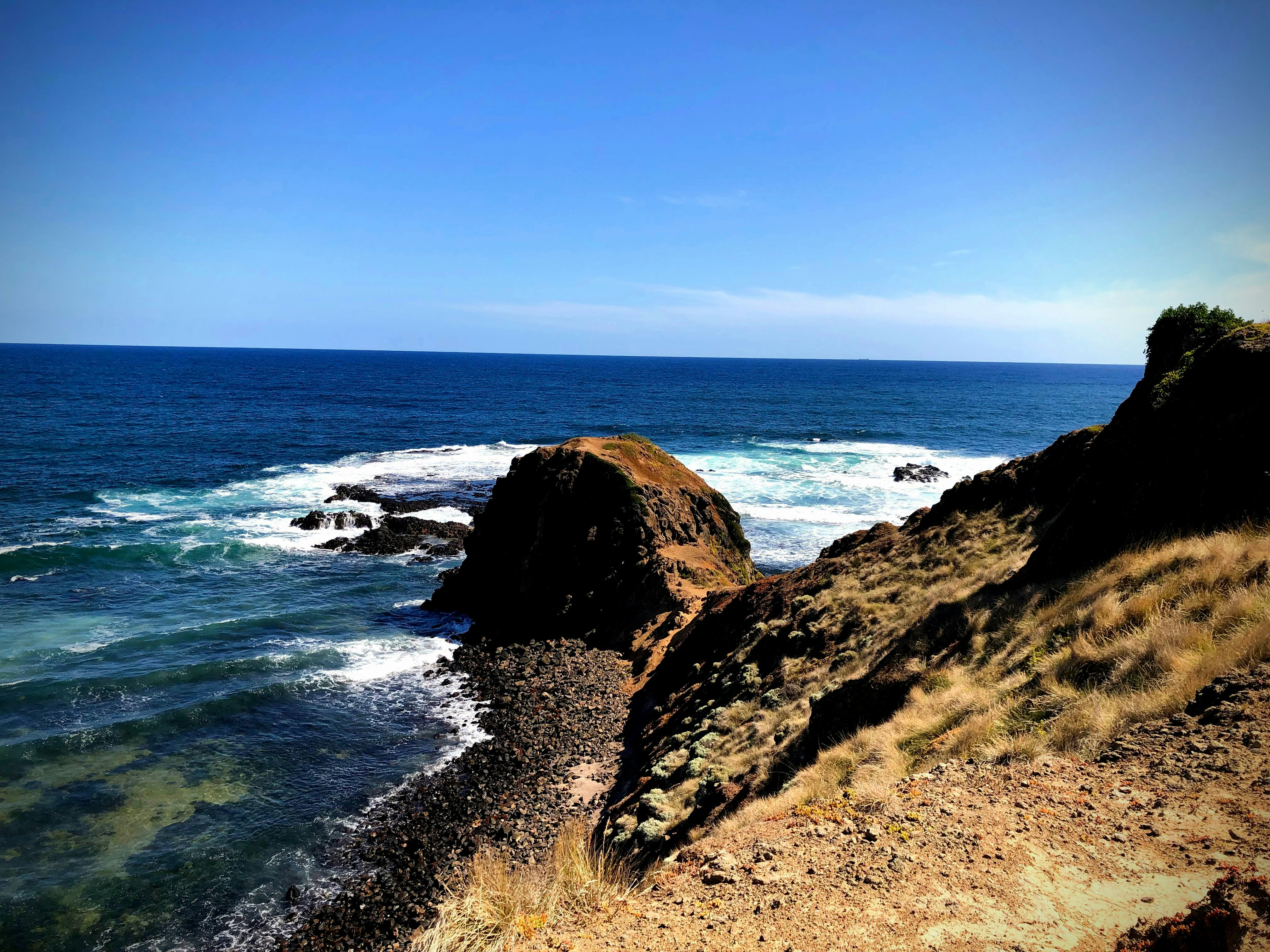 Waves crash against rocky cliffs under a clear blue sky, showcasing the dynamic interaction between land and ocean. The rugged terrain is dotted with dry grass and vibrant ocean hues.