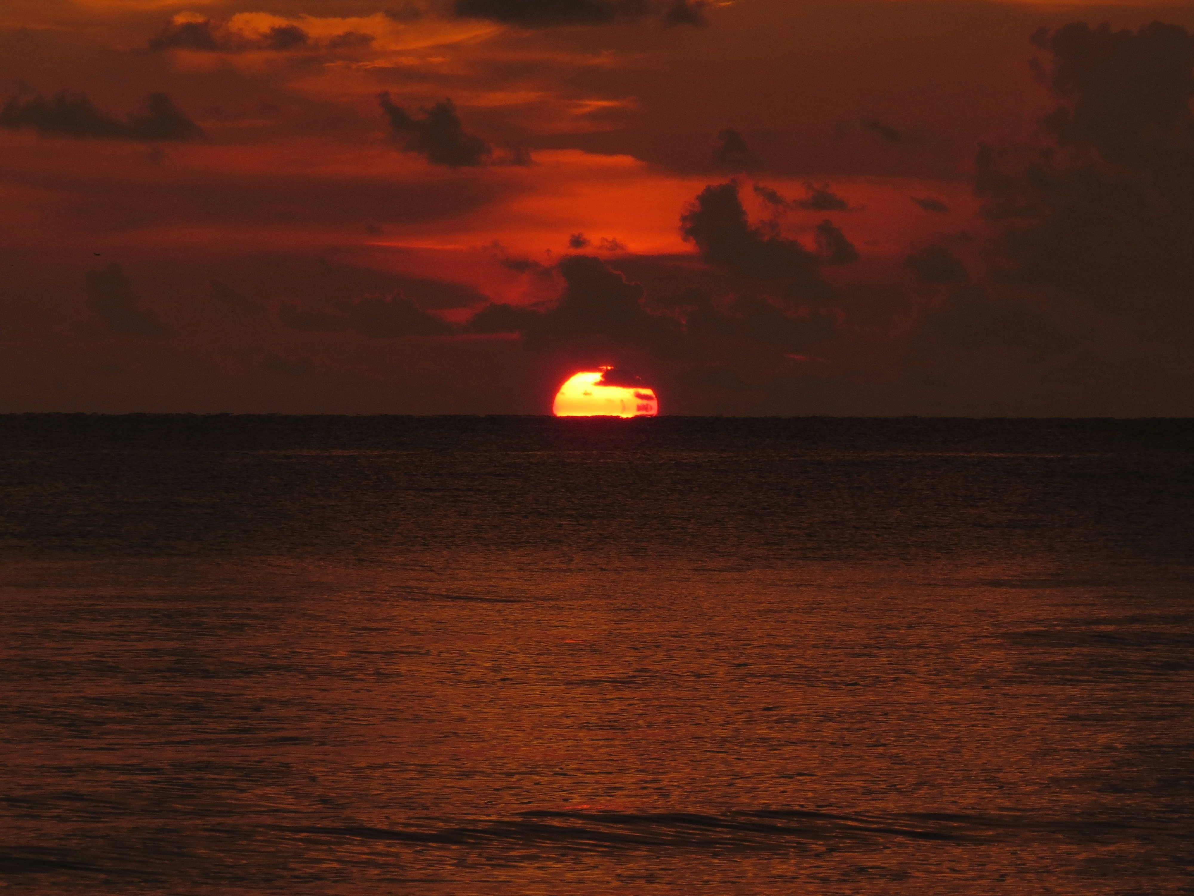 Vibrant sunset casting warm hues over the ocean, with the sun partially submerged on the horizon. Dark clouds frame the scene, enhancing the dramatic effect.