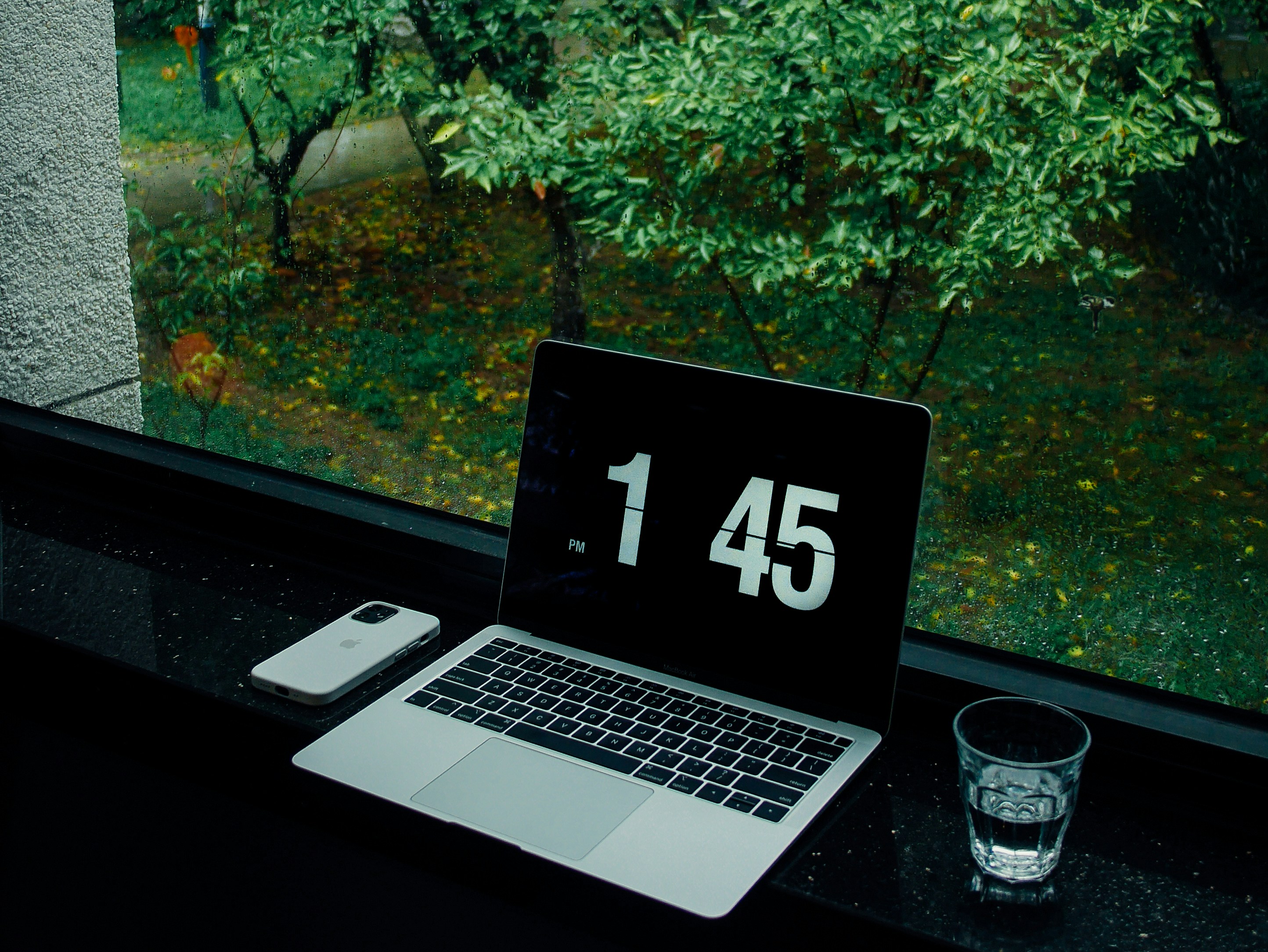 Silvery laptop on a dark windowsill displays a large 1:45 readout, flanked by a smartphone and a glass. Rain-soaked greenery visible through the window sets a moody backdrop for this minimal desk setup.