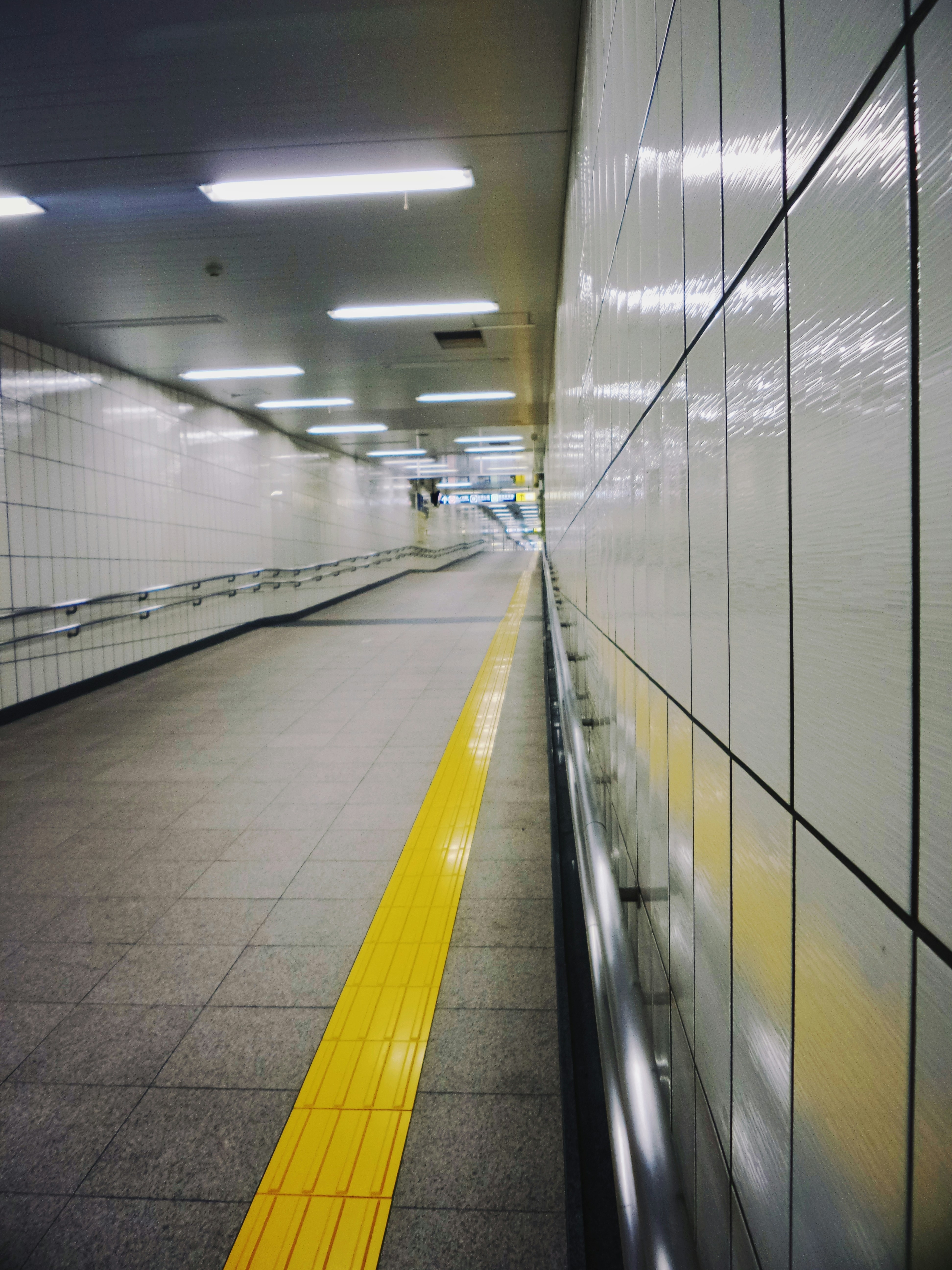A well-lit subway corridor featuring smooth tiles and a prominent yellow guiding line along the floor.