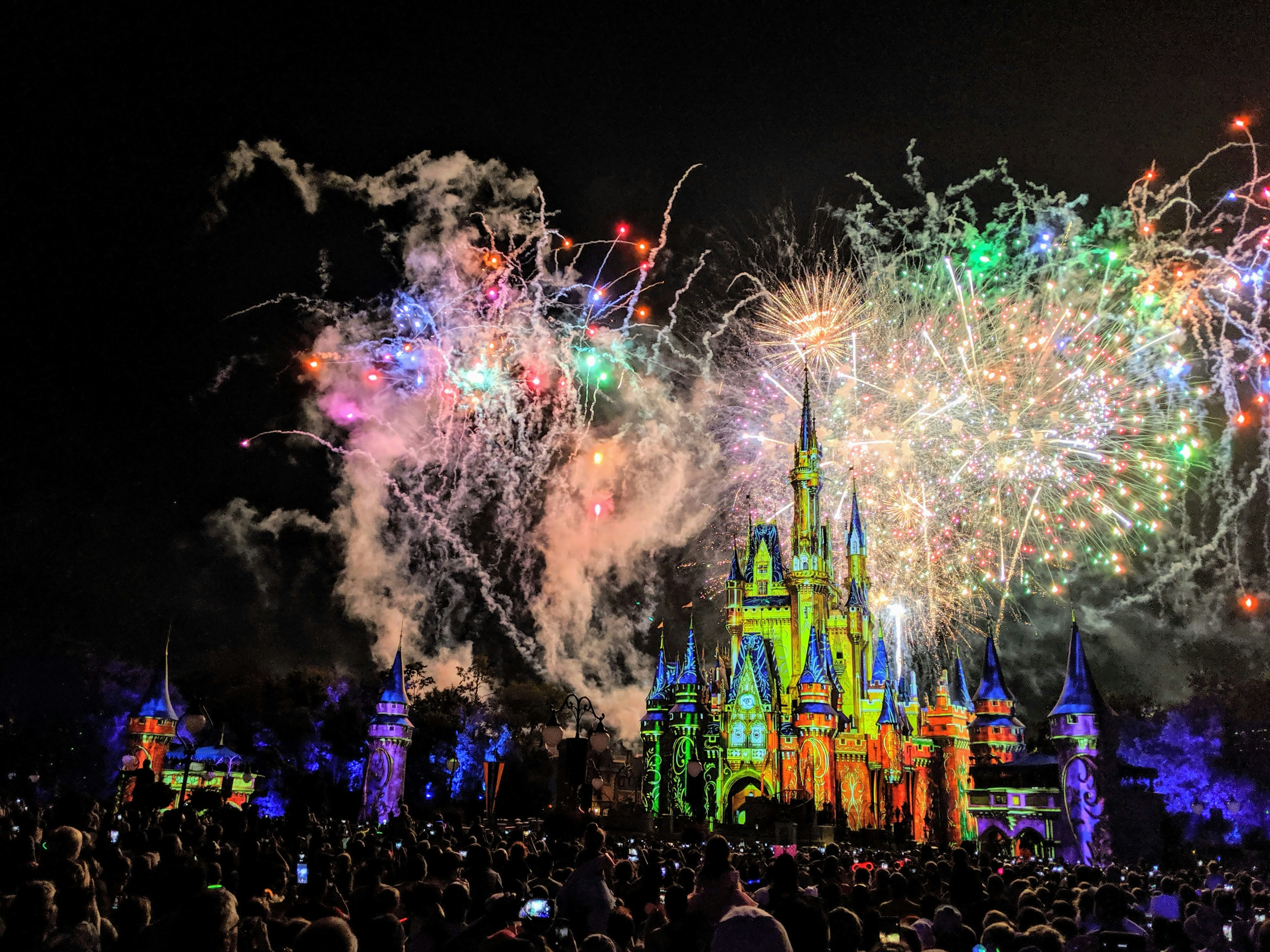 Fireworks erupt above a vibrant castle, illuminating the night sky and captivating a crowd below. The scene is enhanced by colorful projections on the castle's facade.