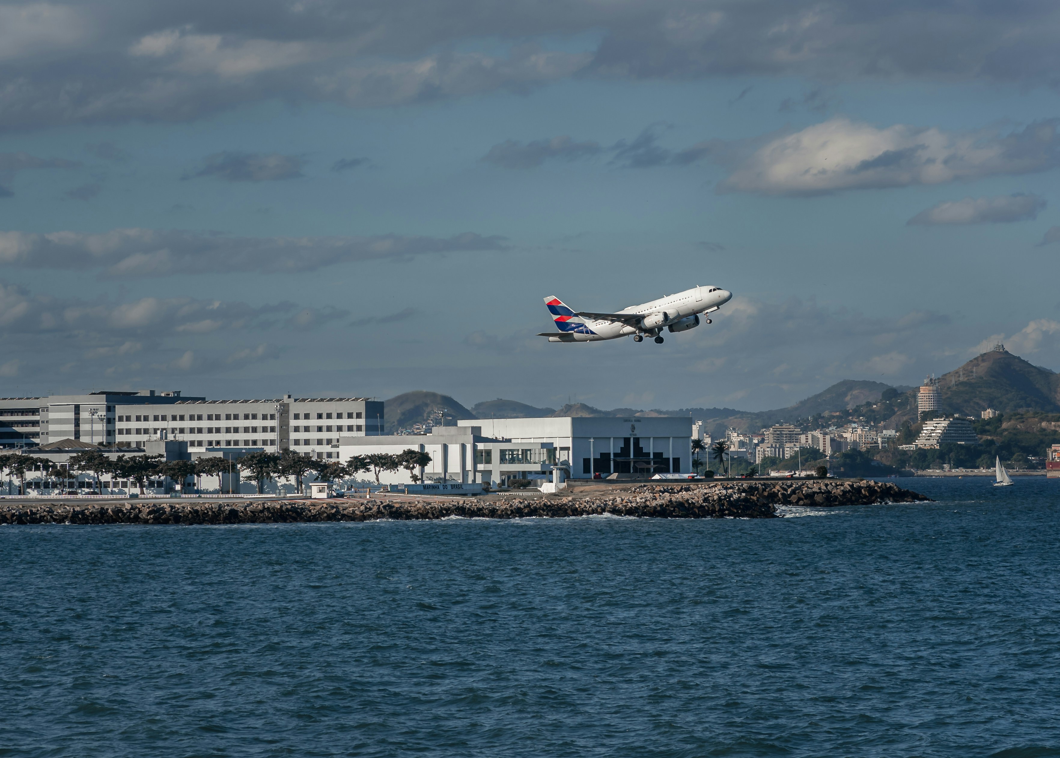 Airplane taking off over a coastal city with mountains in the background under a partly cloudy sky.