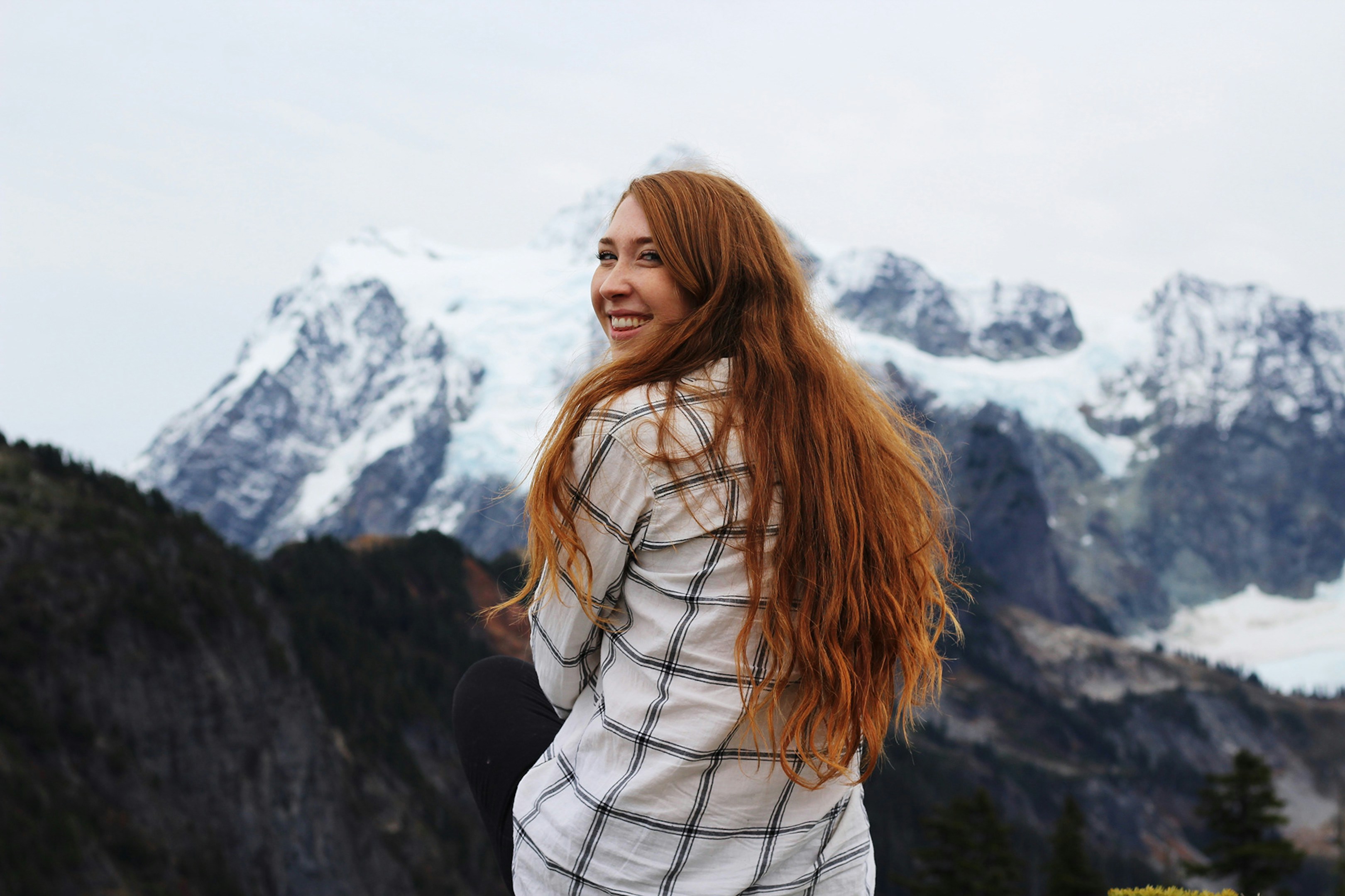 Person with long hair smiling against a backdrop of snow-capped mountains.