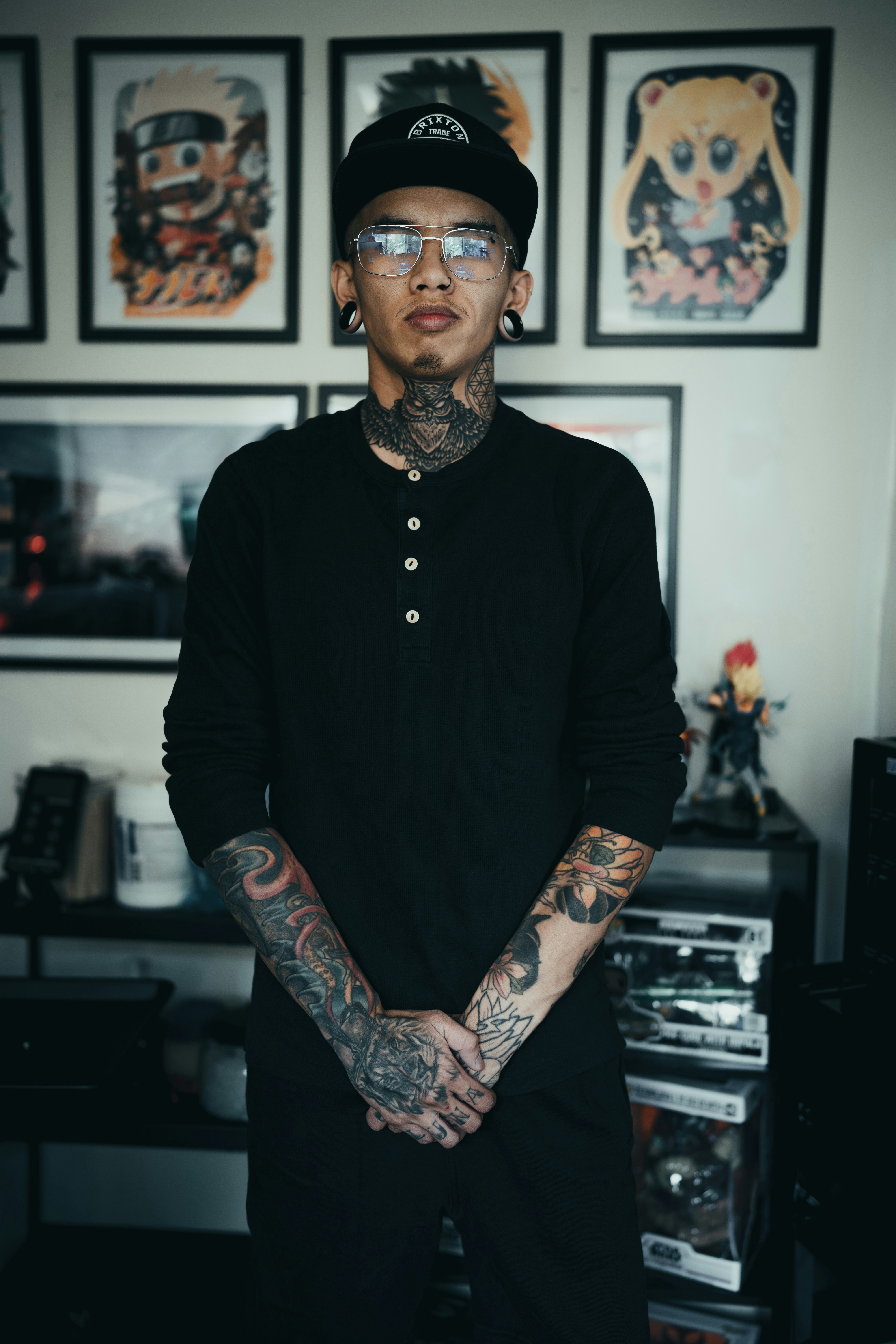 Tattooed individual stands confidently in a room adorned with vibrant artwork. The blend of personal style and creative surroundings highlights a unique cultural aesthetic.