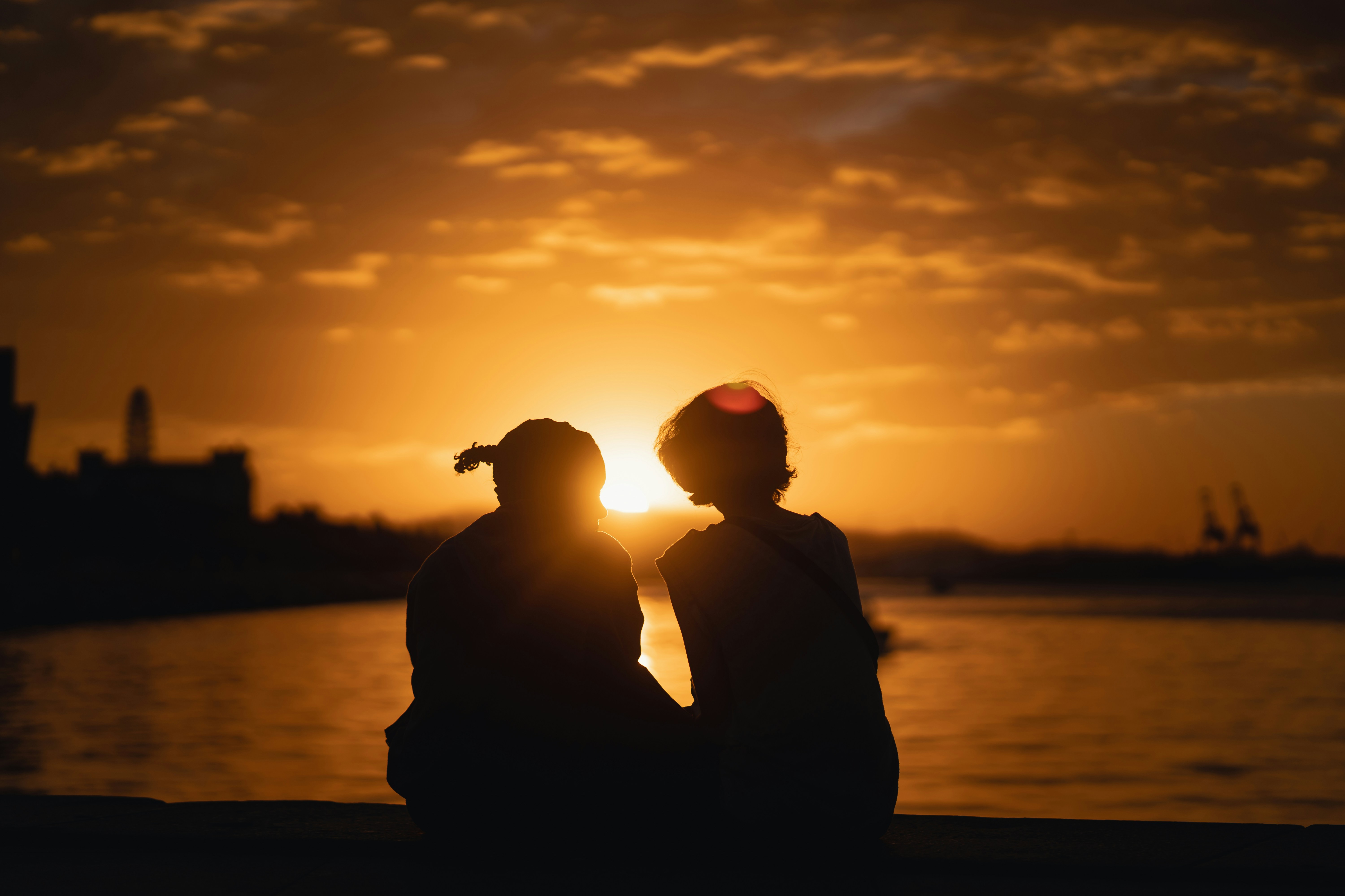 Silhouetted figures sit by a waterfront, framed by a vivid orange sunset sky.