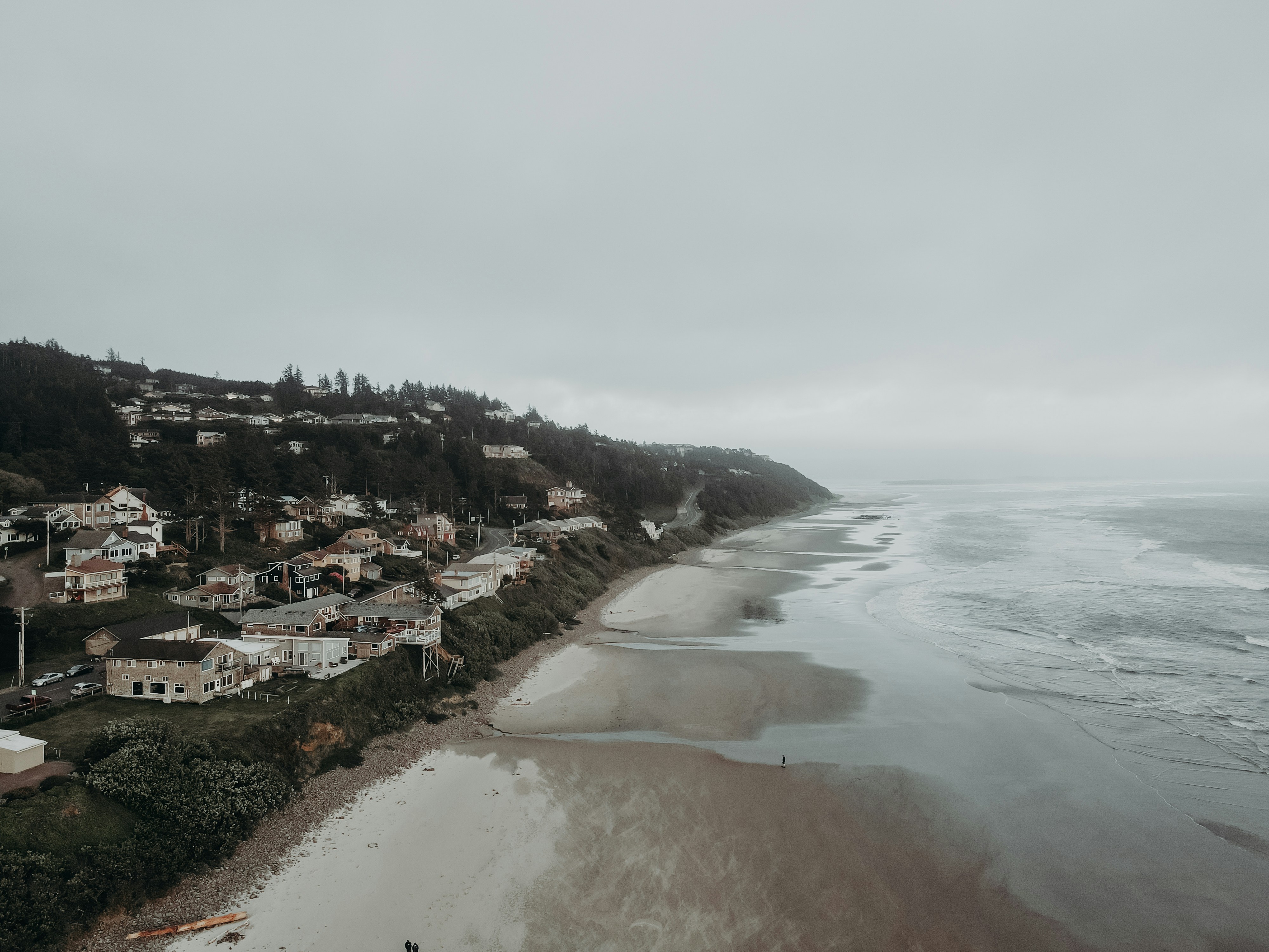 Aerial view of a coastal town nestled against a sandy beach, with gentle waves lapping at the shore under overcast skies.