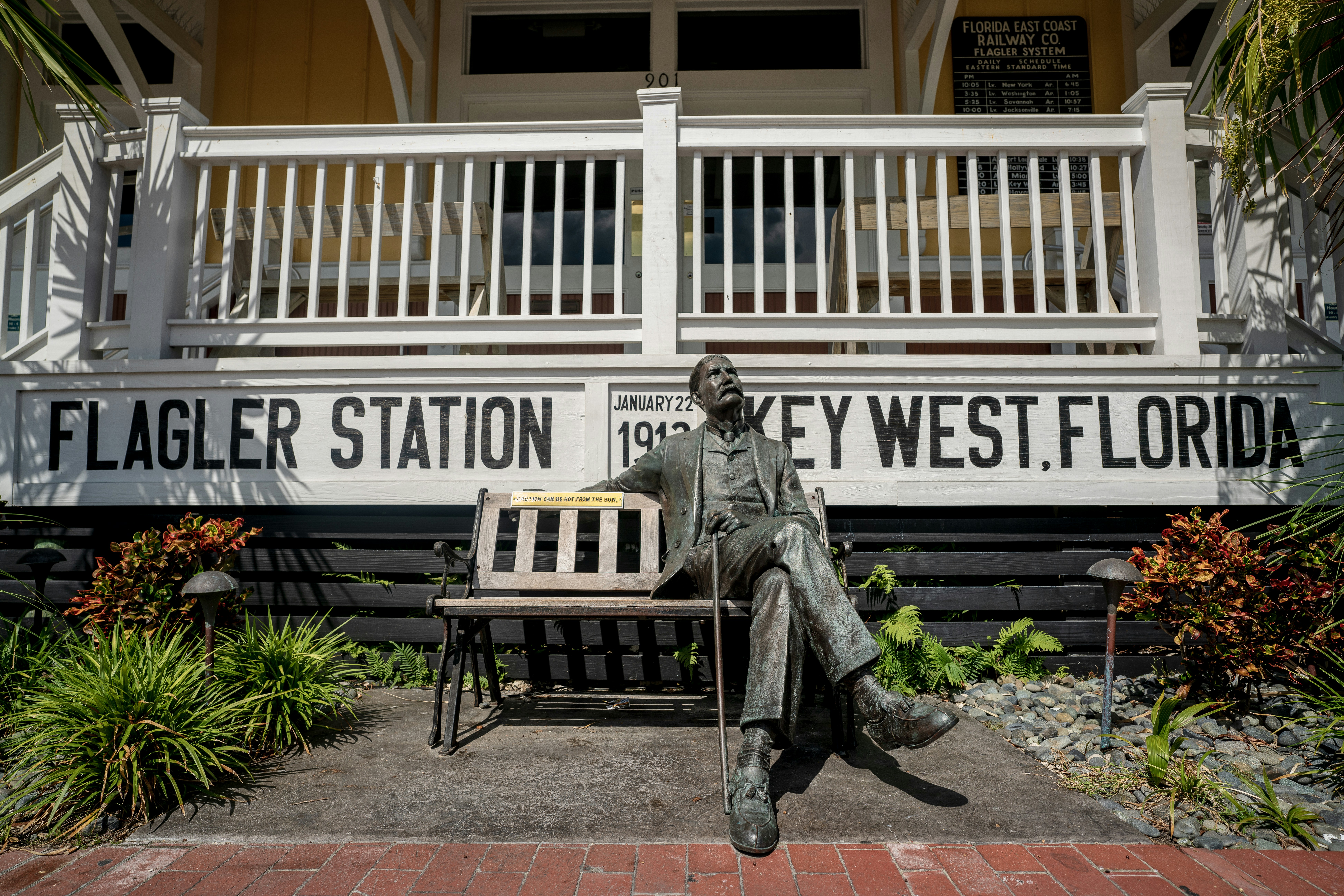 Bronze statue seated on a bench at Flagler Station, Key West, Florida, with a historic sign in the background.