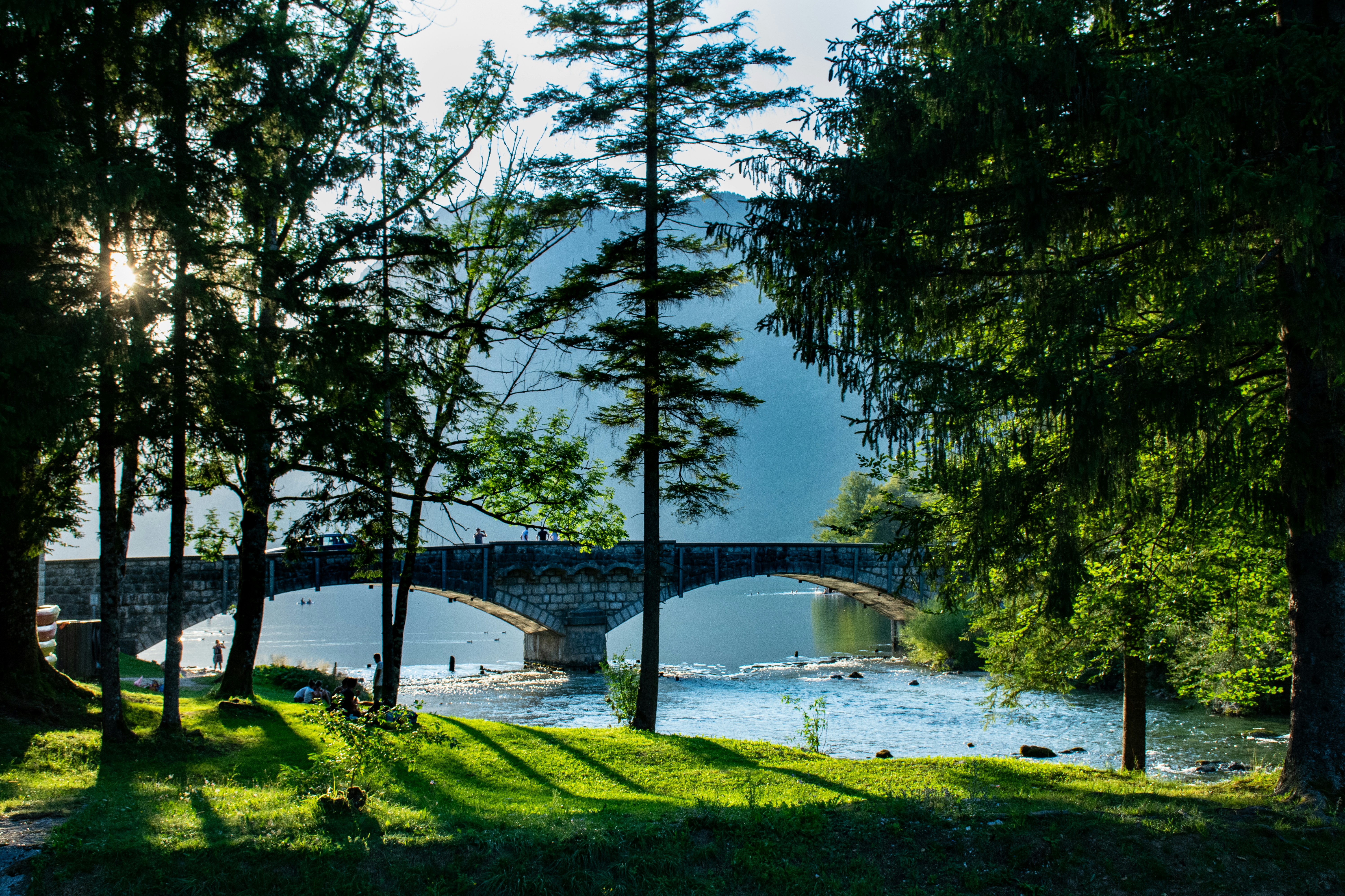 A stone bridge arches gracefully over a shimmering river, framed by lush green trees and dappled sunlight. The serene landscape invites a moment of reflection.