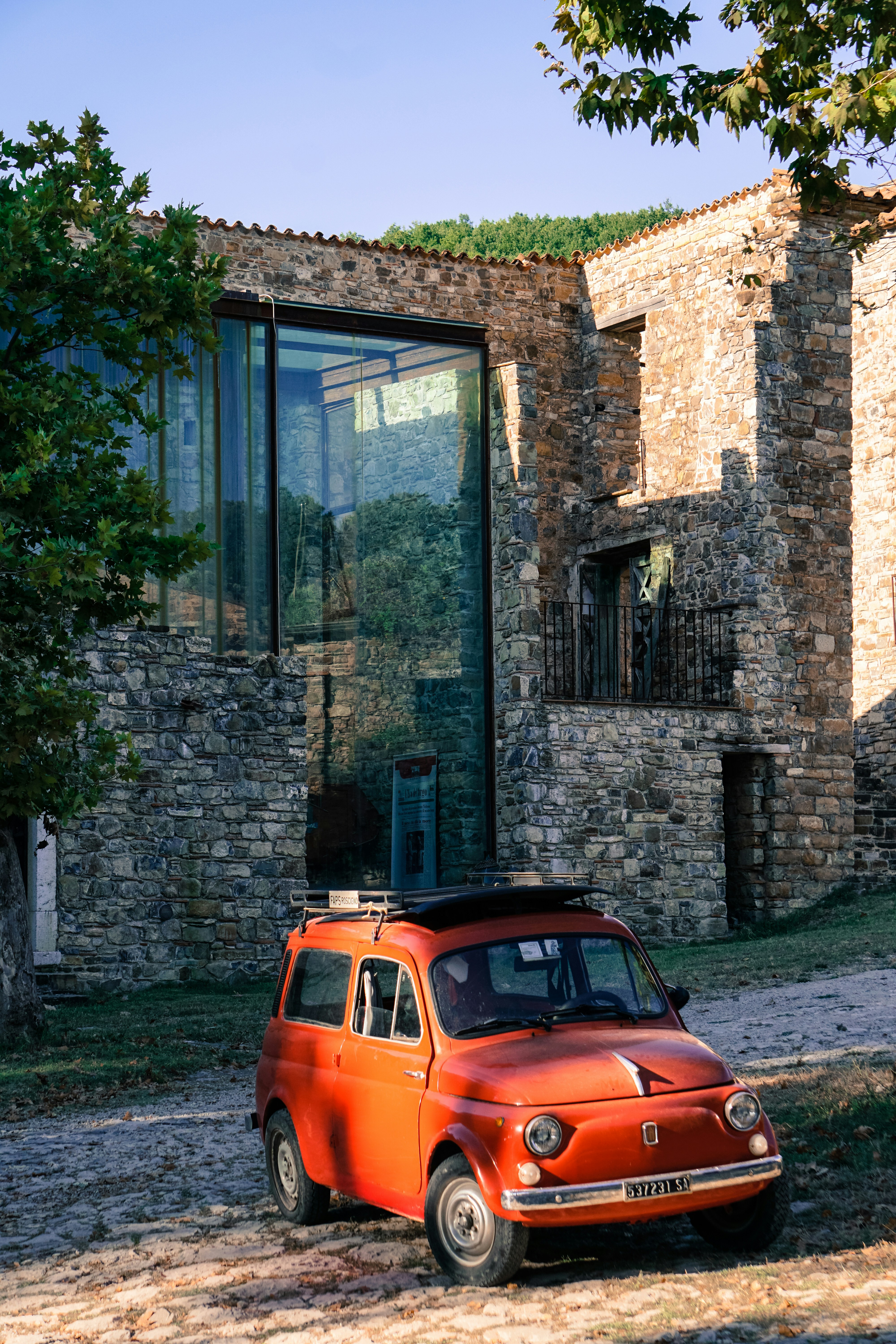 Classic orange car parked in front of a contemporary glass and stone building, surrounded by greenery.