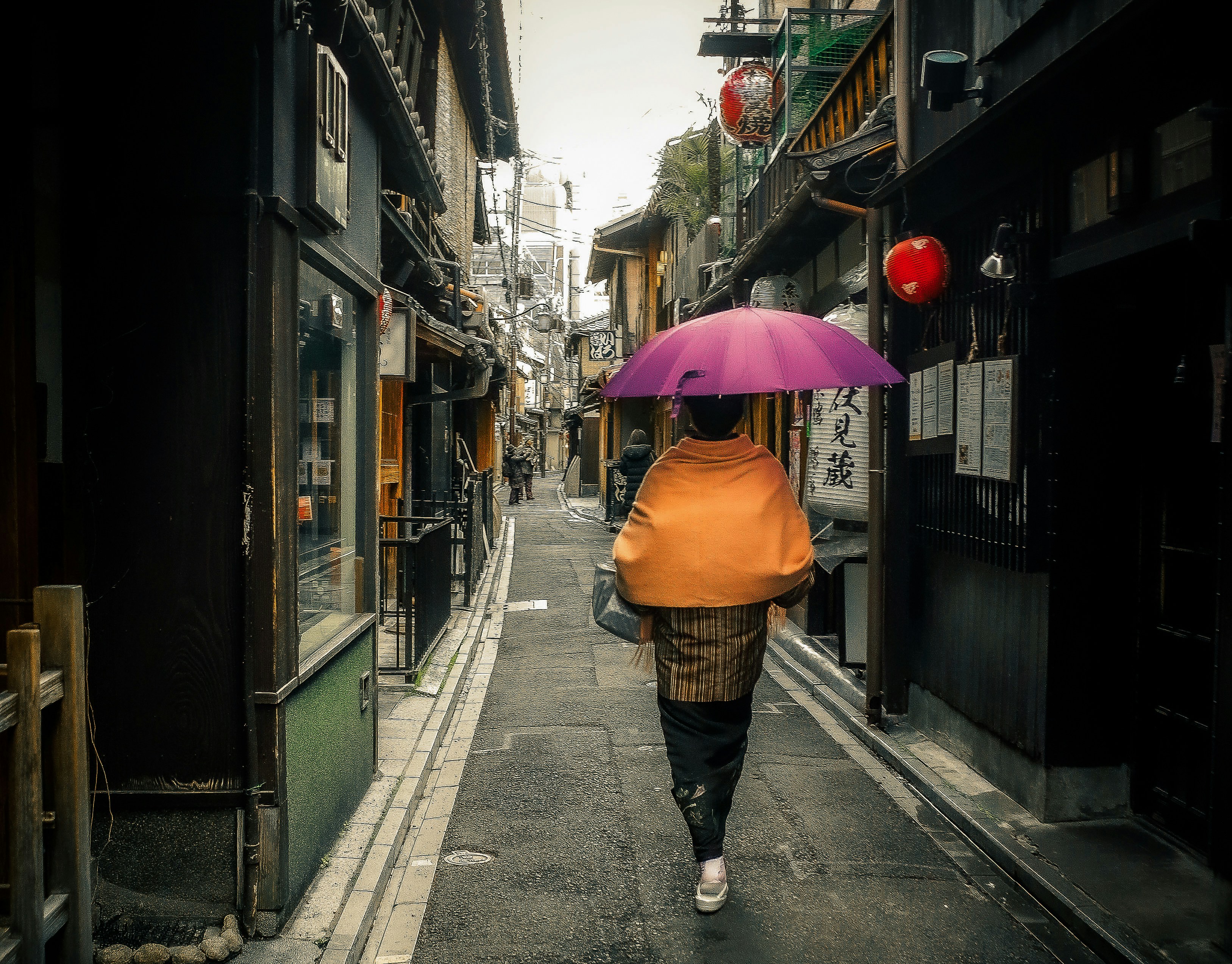A person in an orange coat walks down a lantern-lit alley, holding a purple umbrella. Traditional storefronts and hanging lanterns frame the wet street, capturing a quiet urban moment.