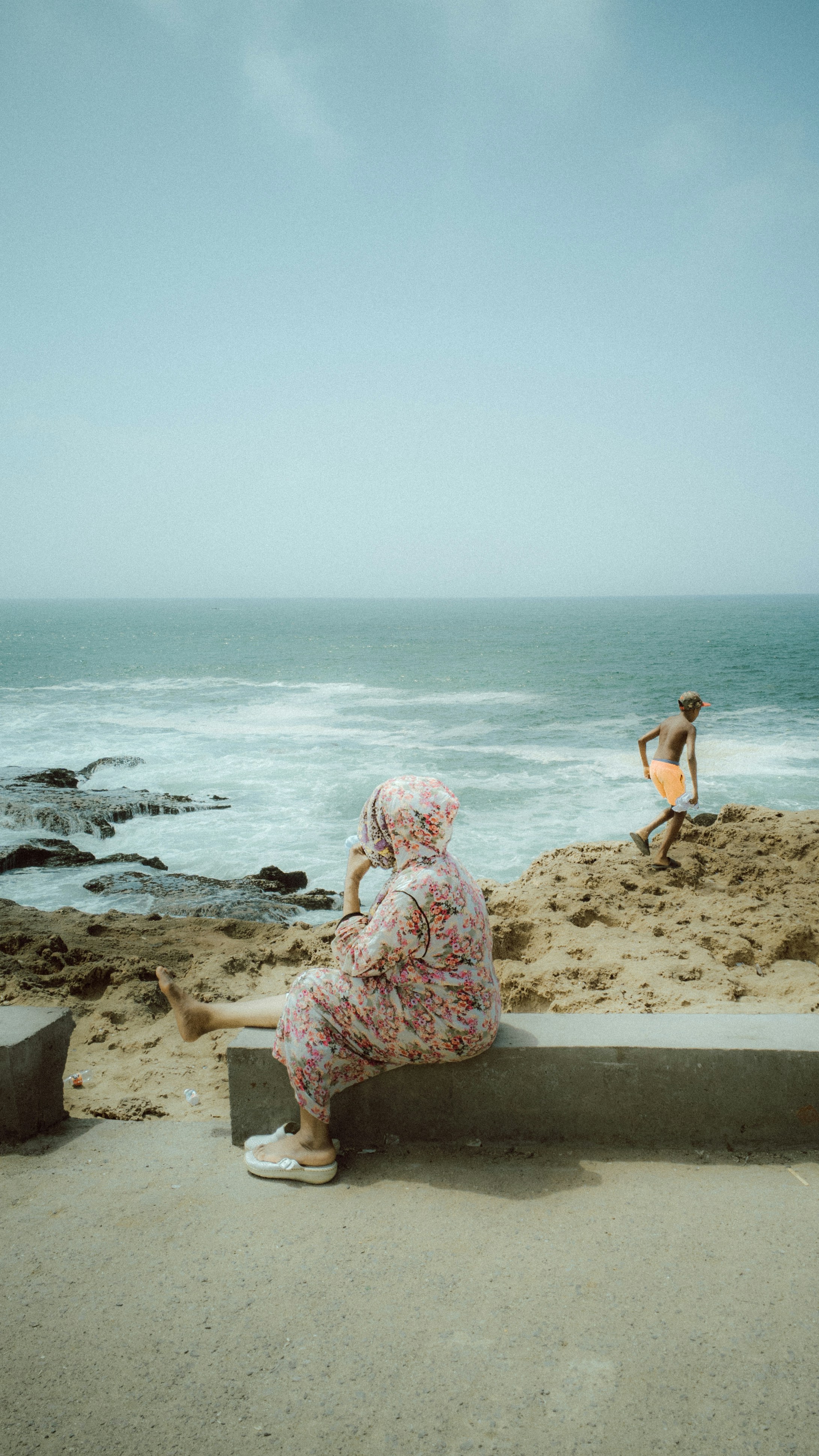 A woman in a floral robe sits contemplatively on a concrete bench, while a boy plays near the waves, capturing a serene moment by the ocean.