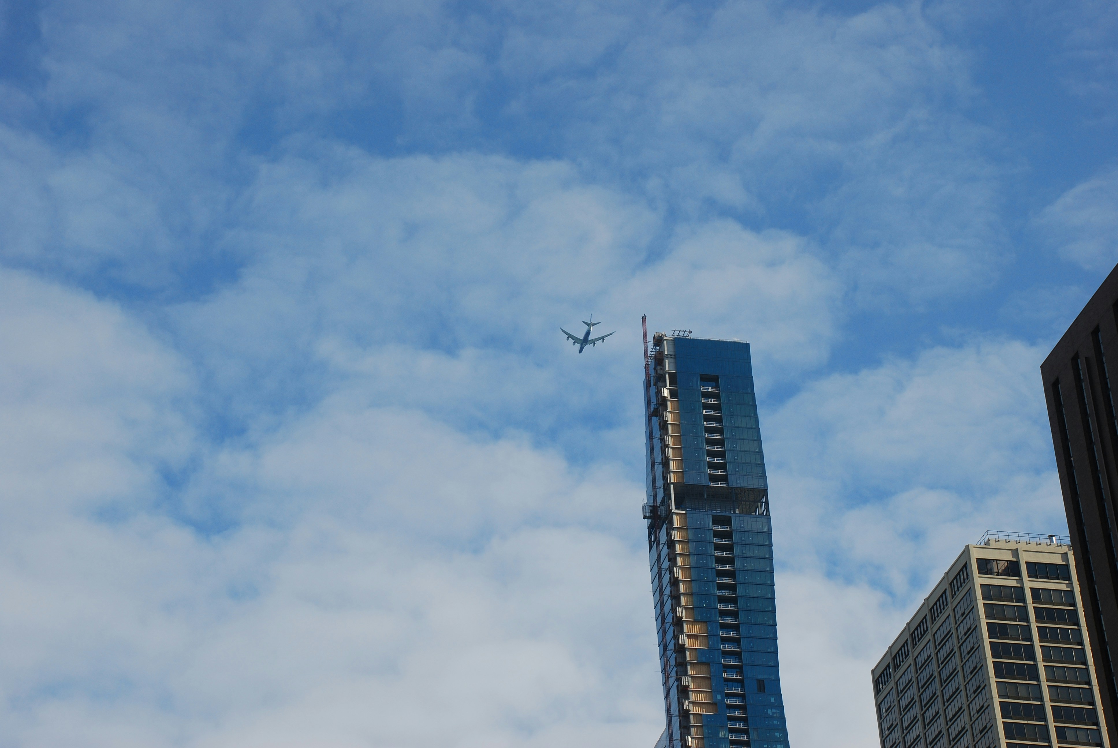 An airplane soars above a modern skyscraper under a partly cloudy sky, highlighting the contrast between architecture and aviation.