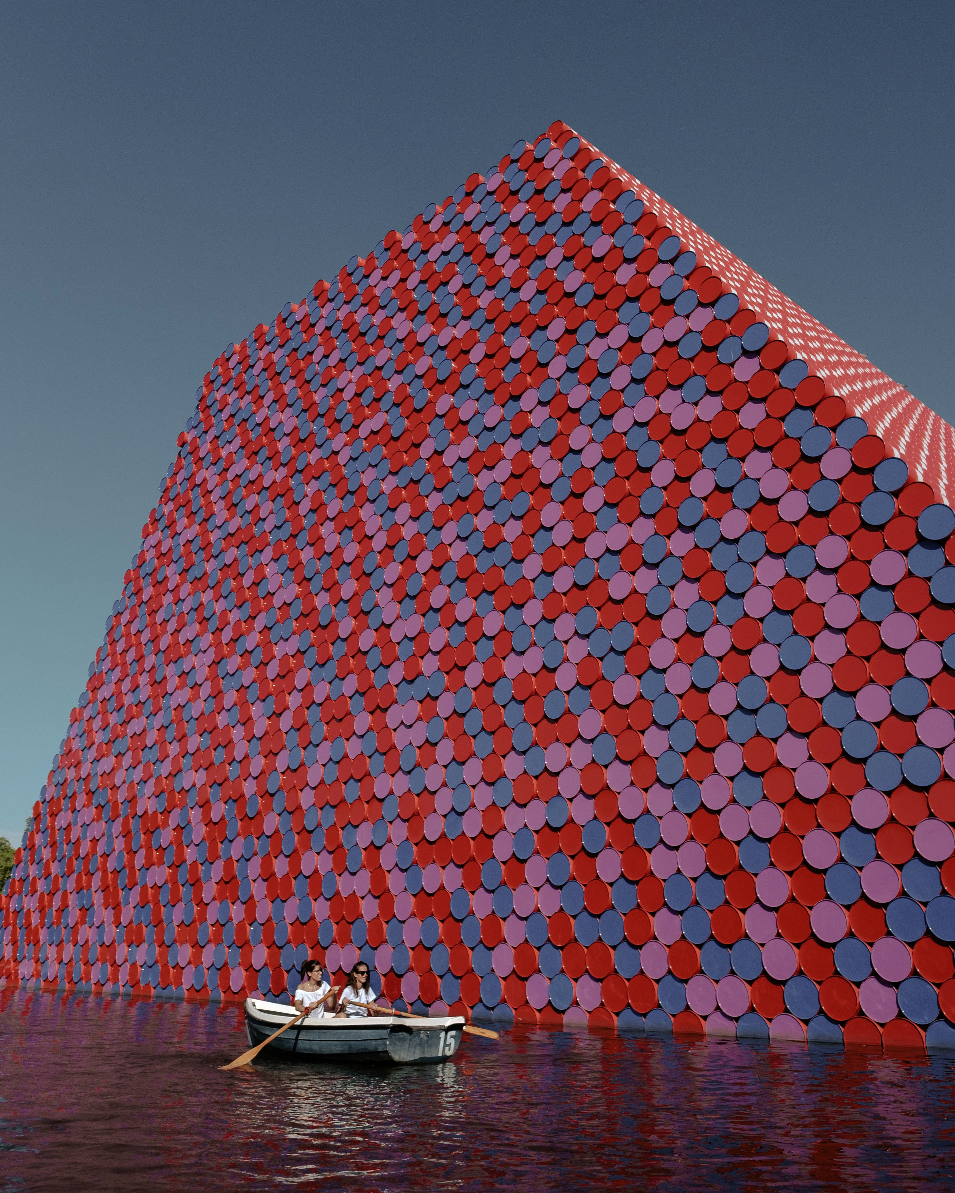 Vibrant geometric structure composed of circular panels rises above the water, while two individuals row in a small boat nearby.