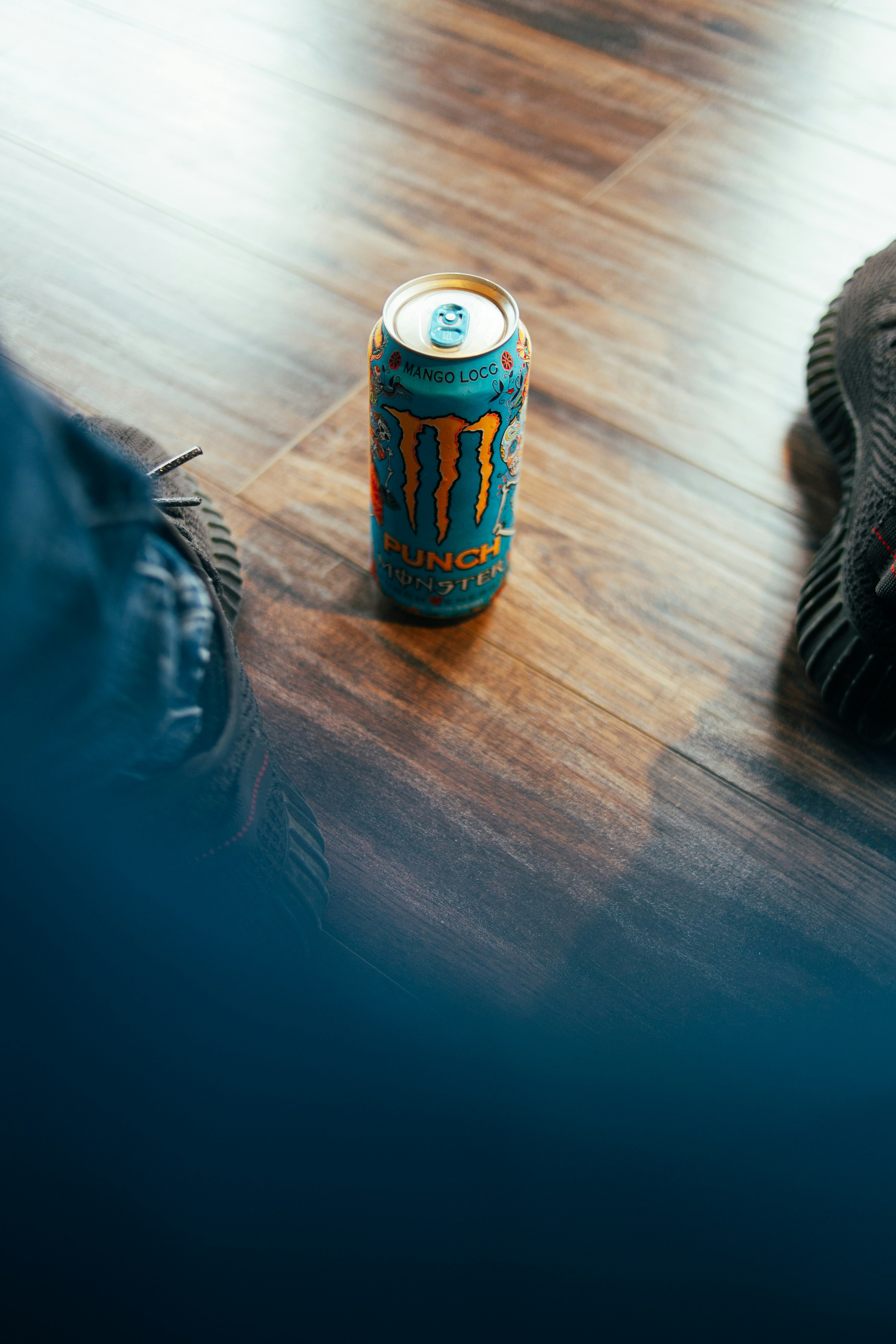 Colorful Monster energy drink can rests on wooden floor, surrounded by casual footwear. The scene captures a moment of relaxation and energy.
