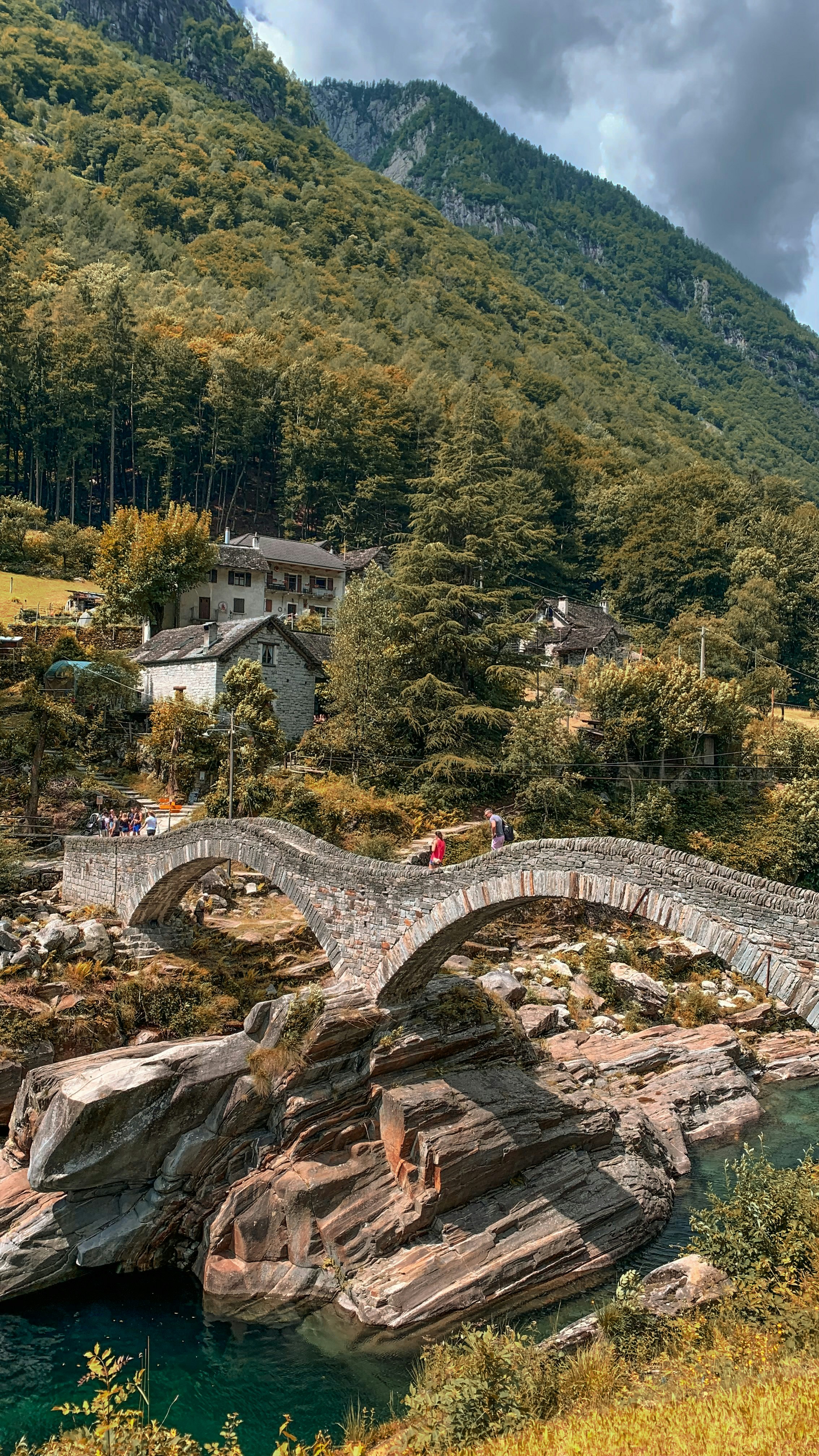 A historic stone bridge gracefully arches over a rocky riverbed, surrounded by lush greenery and quaint houses. The scene captures a moment of tranquility in a picturesque valley.