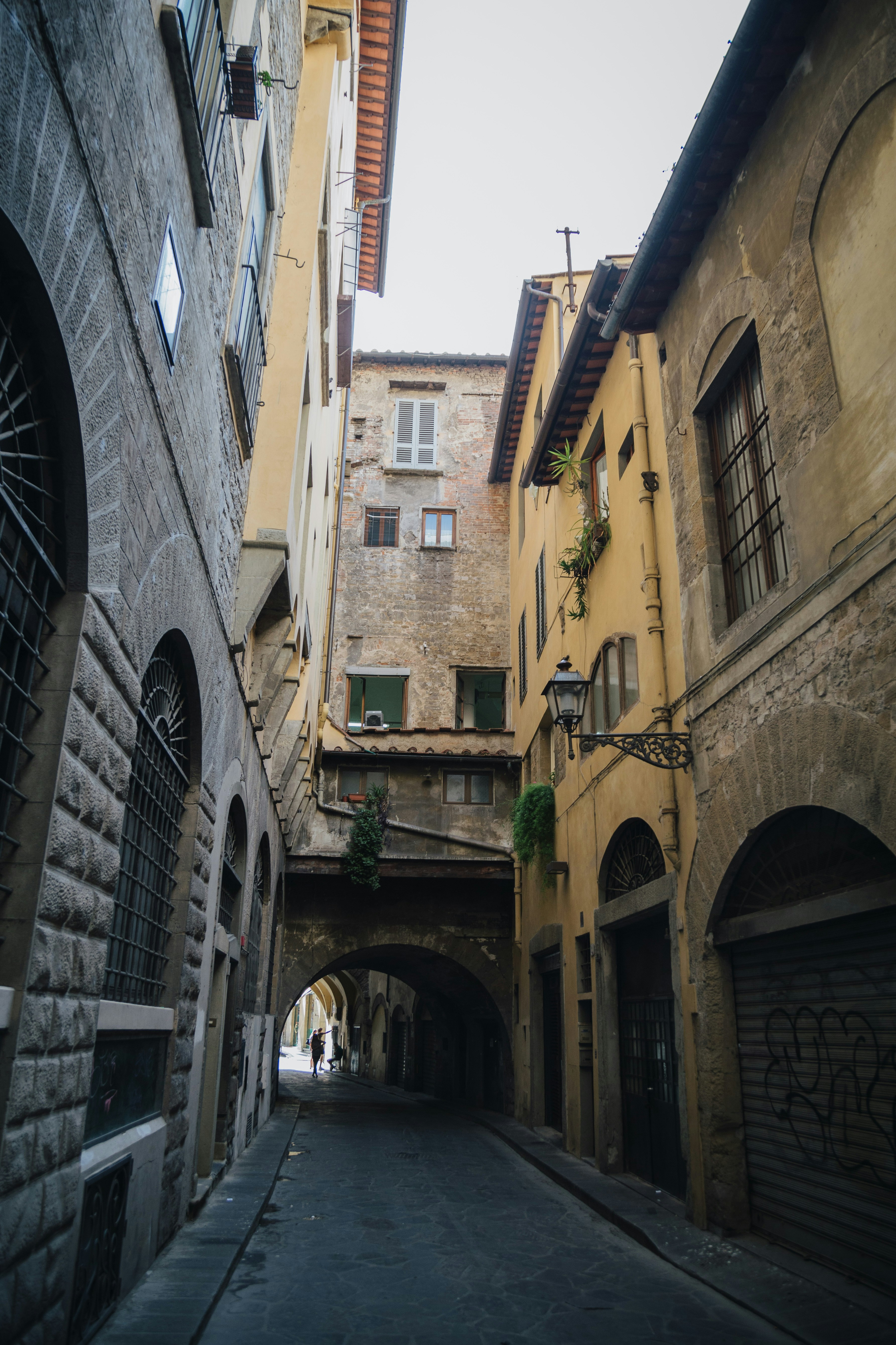 Narrow cobblestone alley flanked by aged buildings with arched passageways and greenery, evoking a sense of timelessness.
