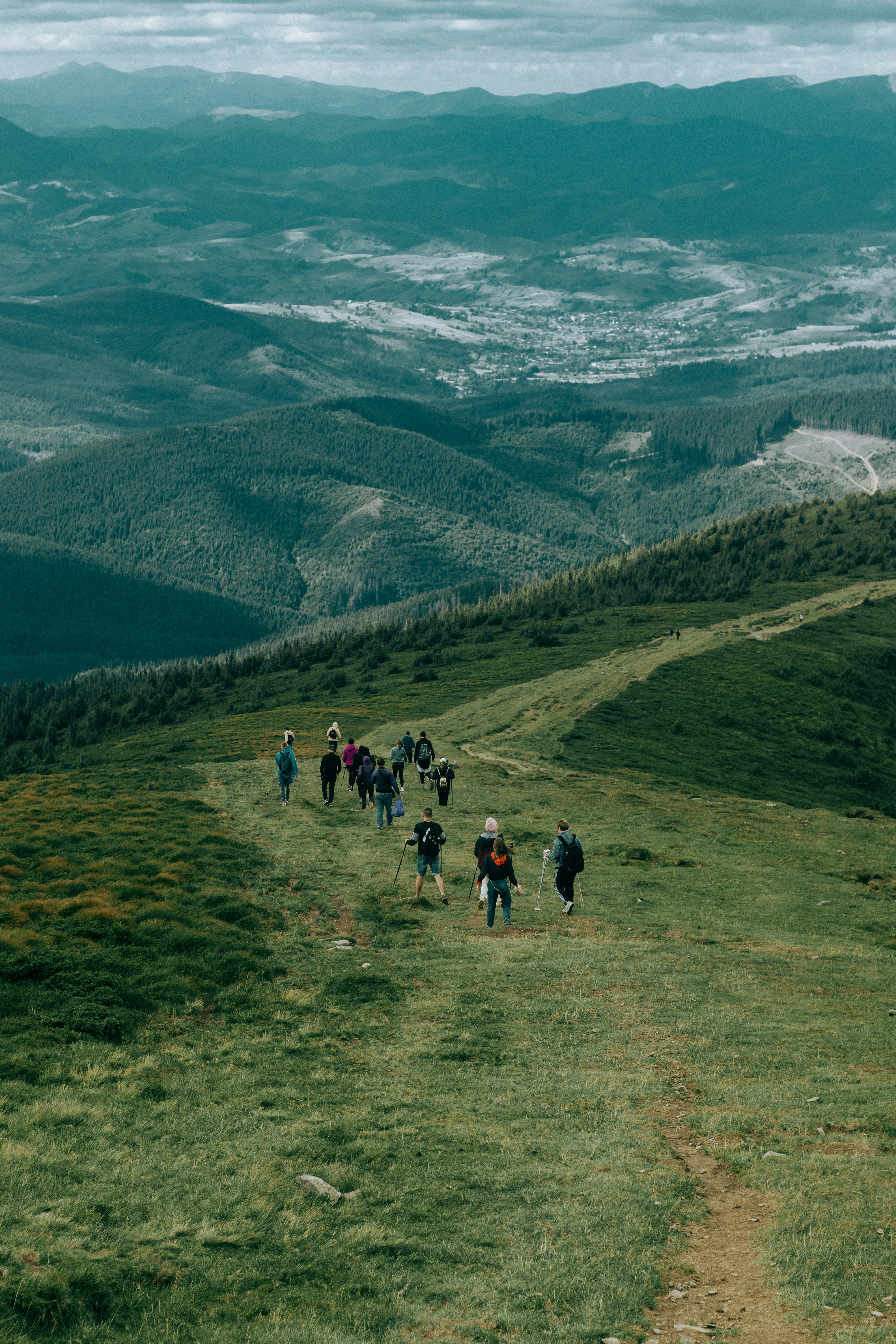 Group of hikers traversing a lush mountain path, surrounded by rolling hills and valleys under a cloudy sky.