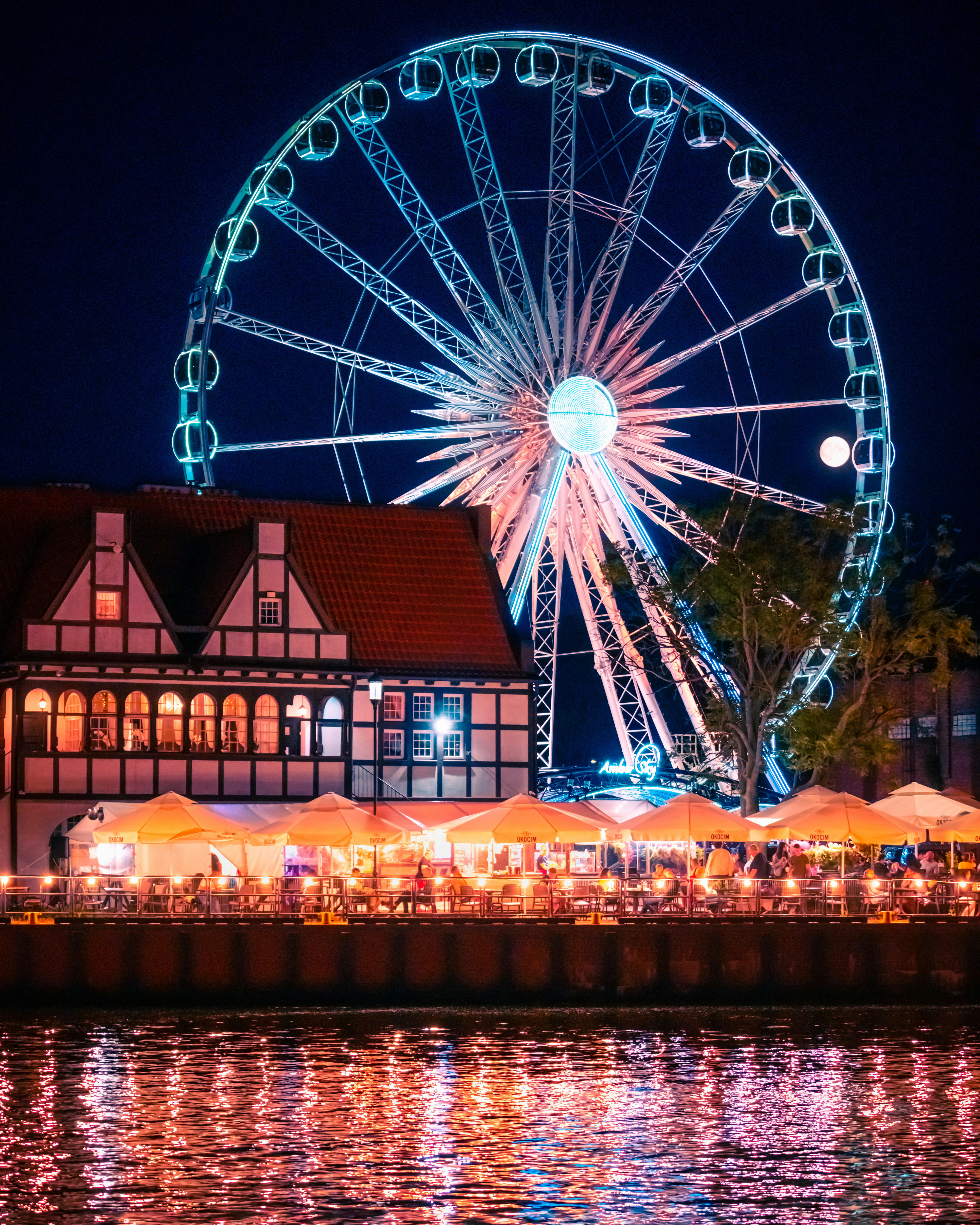 Illuminated Ferris wheel towering over a riverside dining area, reflecting colorful lights on the water's surface at night.