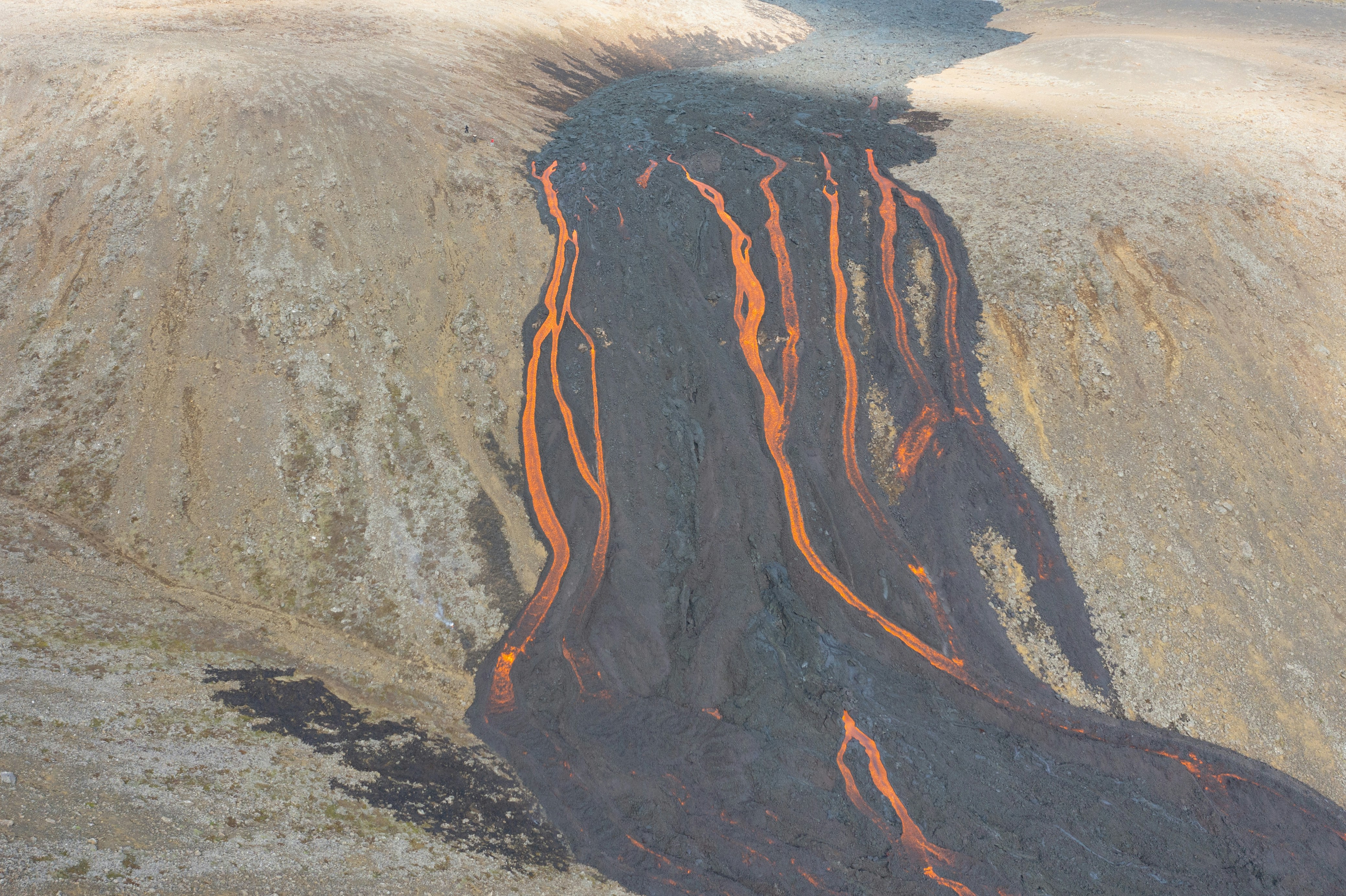 Lava flows cascading down a volcanic landscape, showcasing vibrant orange streaks against dark volcanic rock. The image captures the raw power of nature's geological processes.
