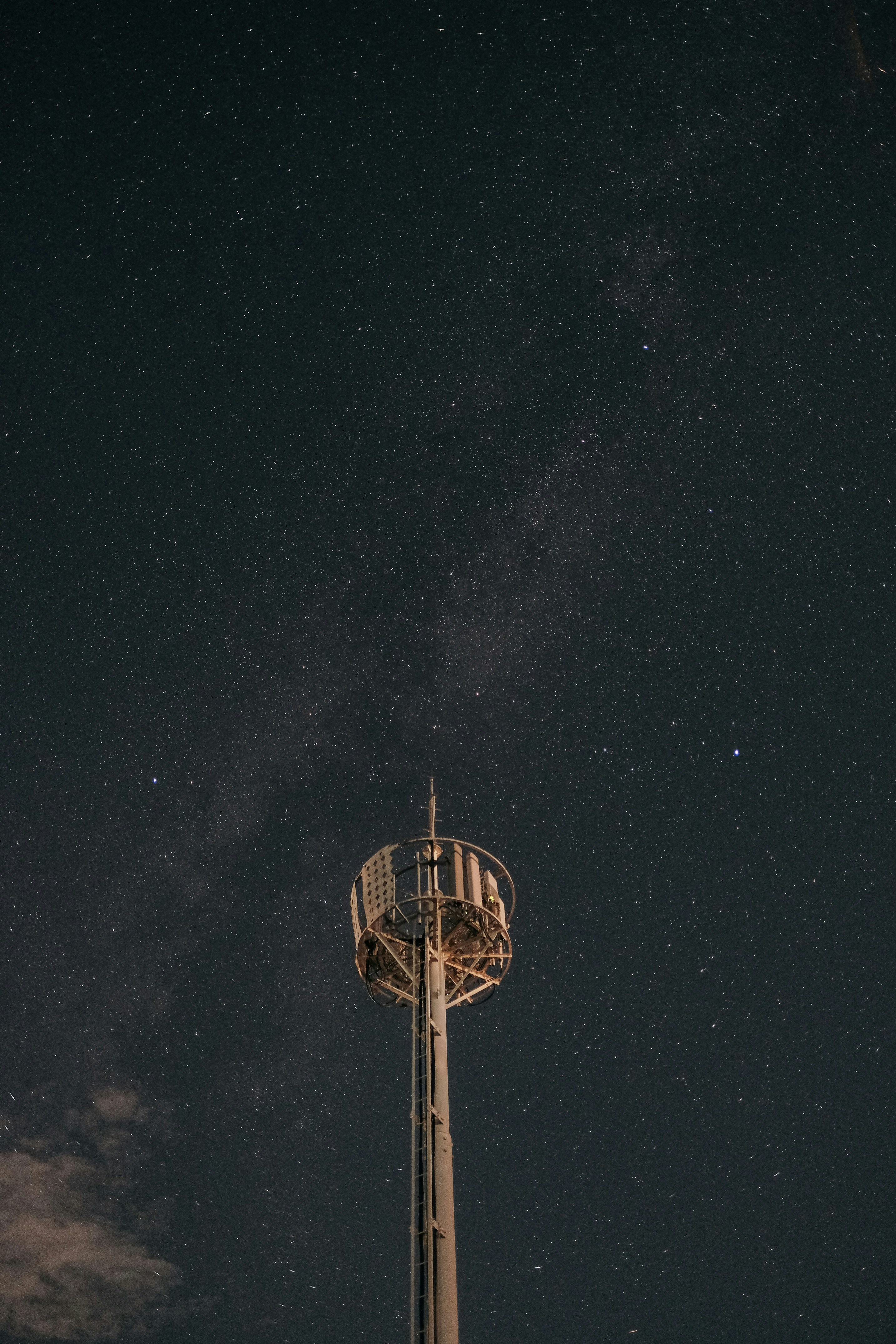 A tall communication tower stands against a star-filled sky, with the Milky Way visible in the background. The scene captures the juxtaposition of human engineering and the vastness of the universe.