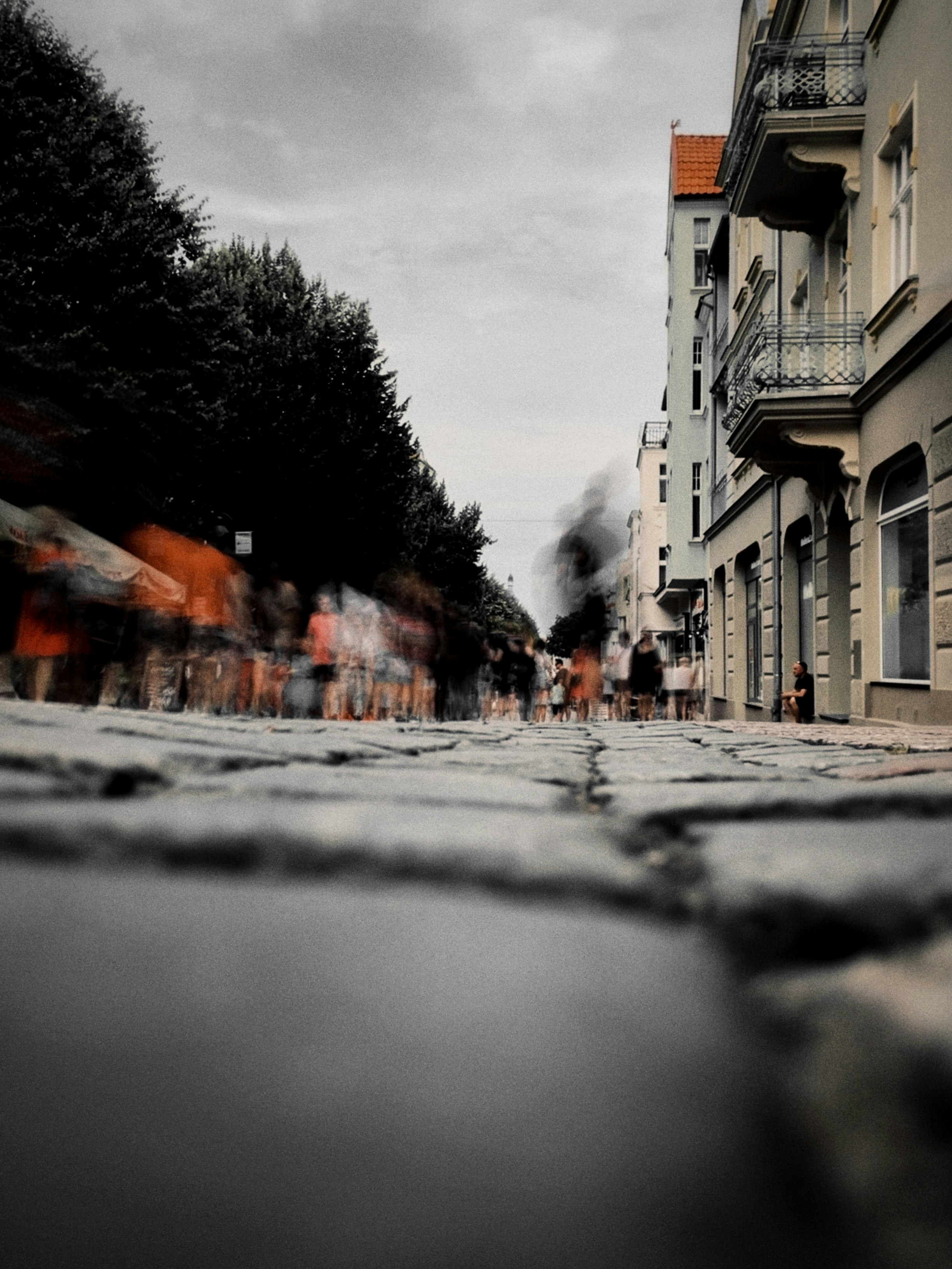 Cobblestone street viewed from a low angle, with blurred figures of pedestrians in motion against a backdrop of buildings and trees.