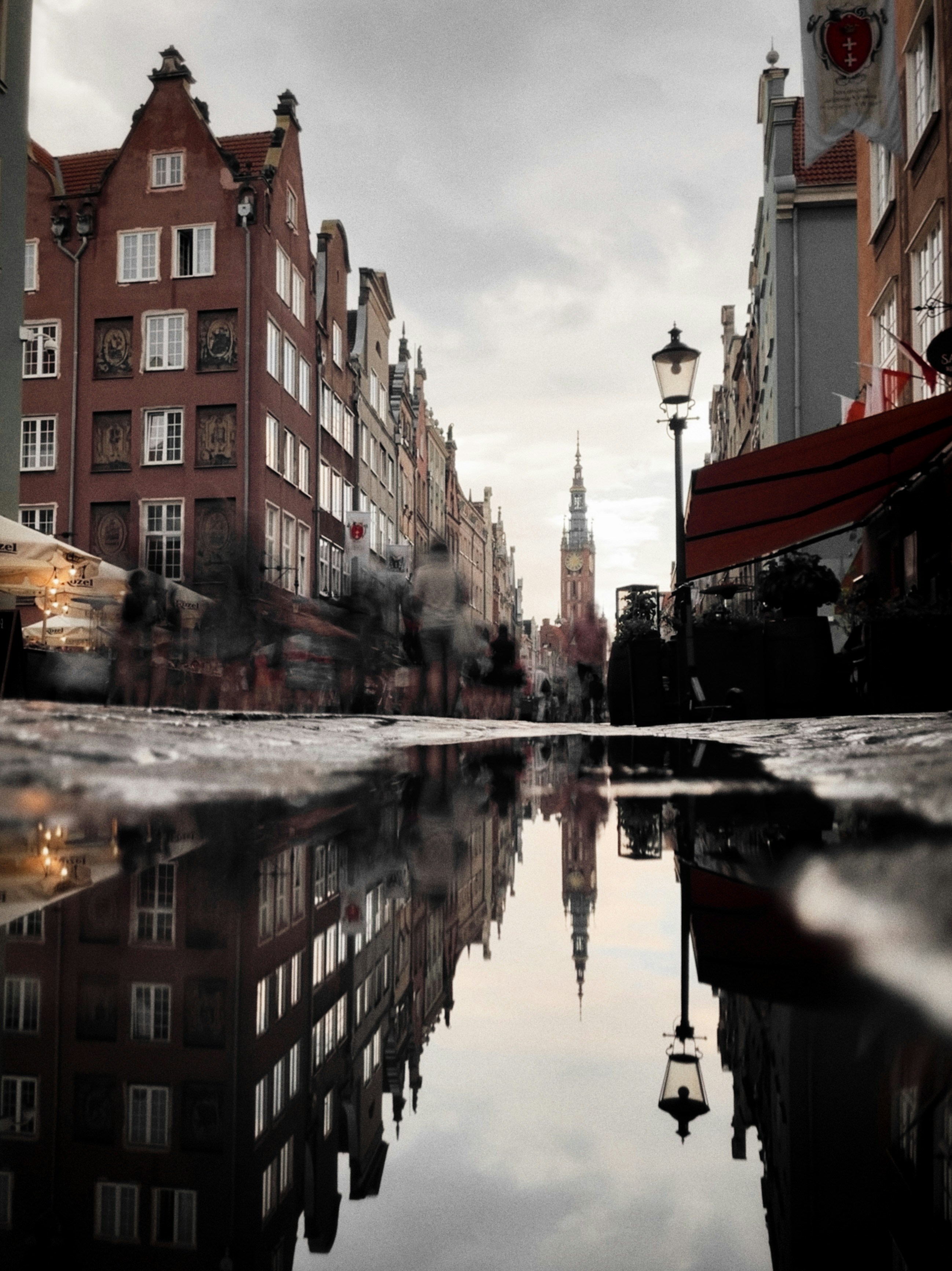 Rain-soaked city street photograph capturing a tall clock tower reflected in a puddle, with red-brick buildings and blurred pedestrians creating a sense of motion.