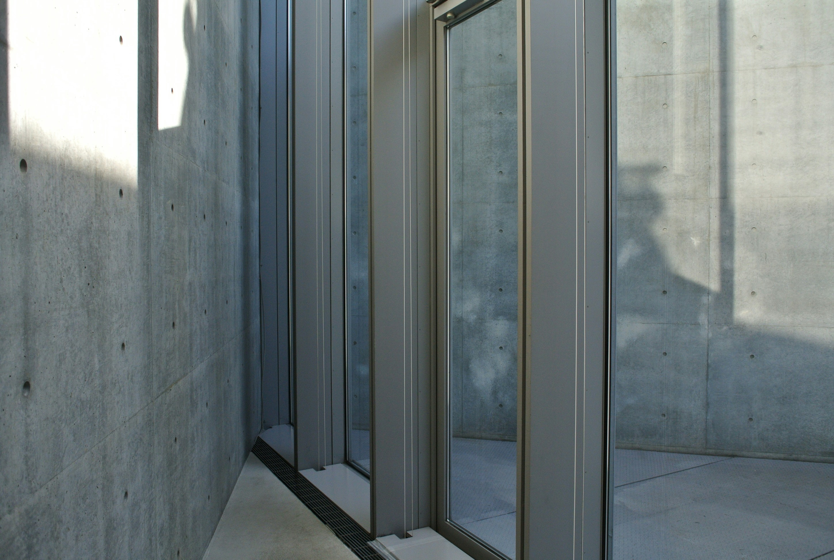 A photograph of a narrow glass corridor between Brutalist concrete walls, featuring tall vertical window frames and soft, directional light.