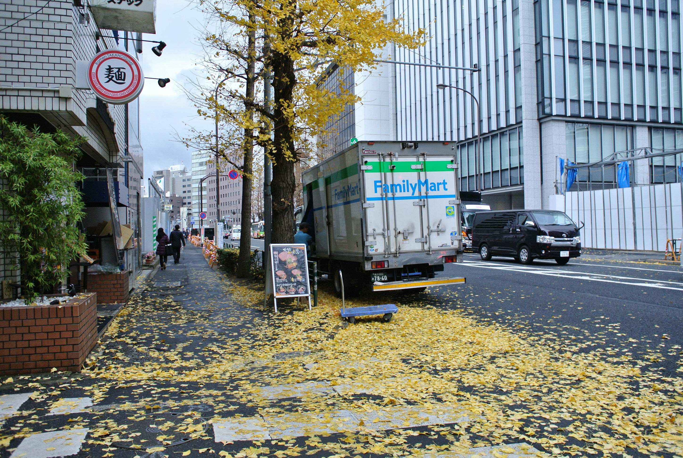 Autumn leaves blanket a city sidewalk as a green FamilyMart delivery truck is parked by a tree, with glass-fronted buildings and street signage along the curb.