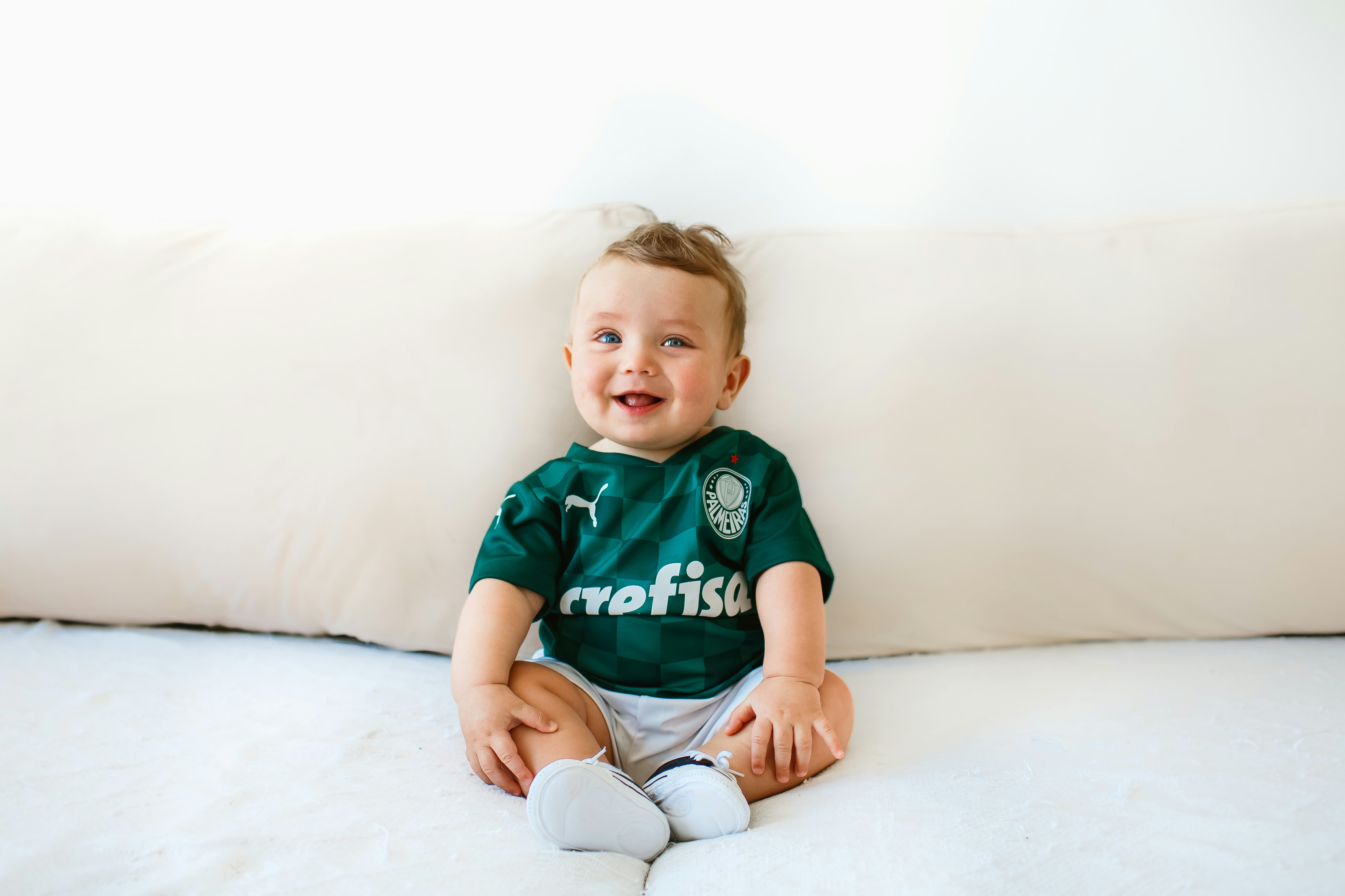 Smiling baby in a green jersey sitting on a white couch, exuding happiness and innocence.