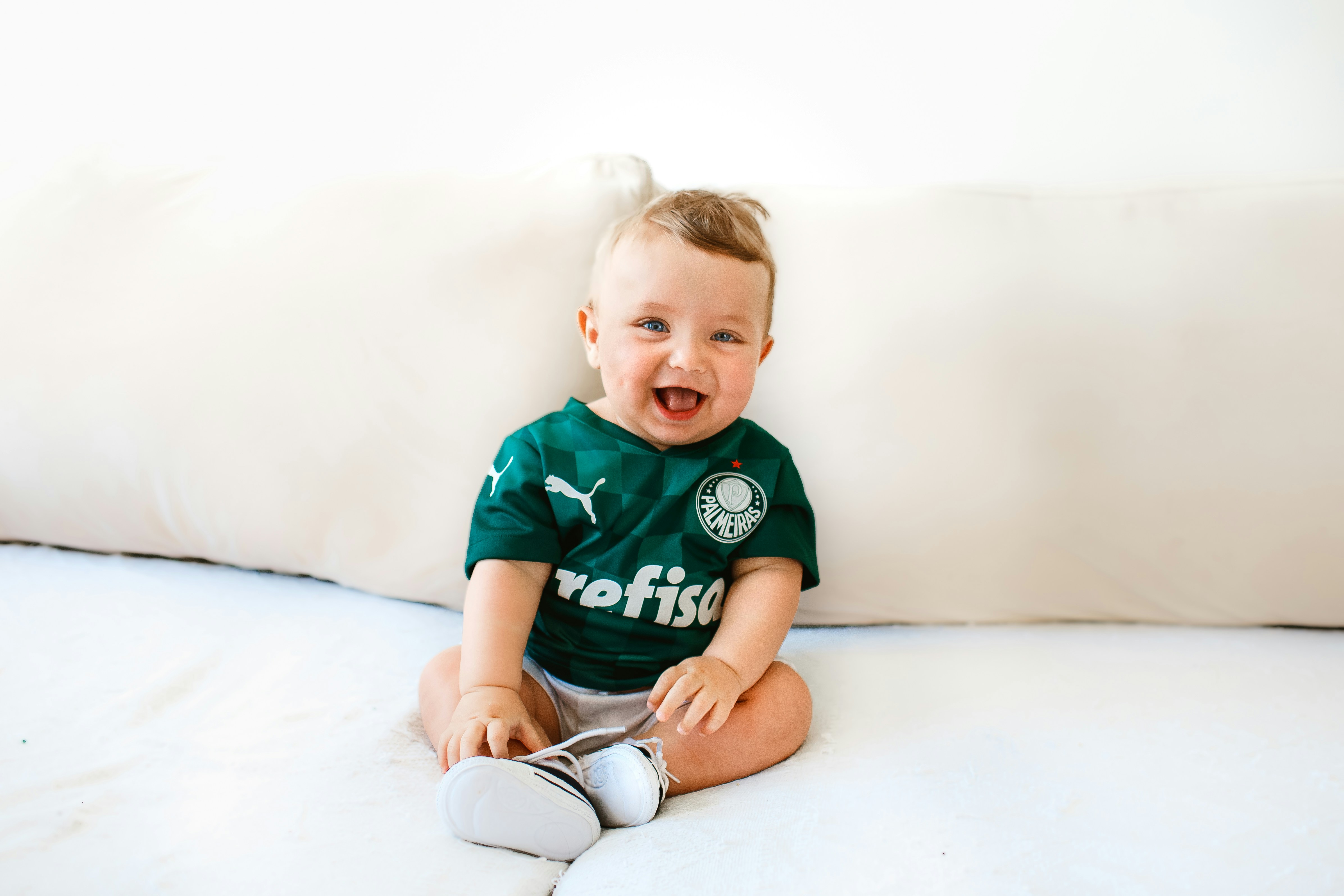Smiling baby dressed in a green sports jersey sitting on a light-colored sofa. The playful expression captures the essence of childhood joy.