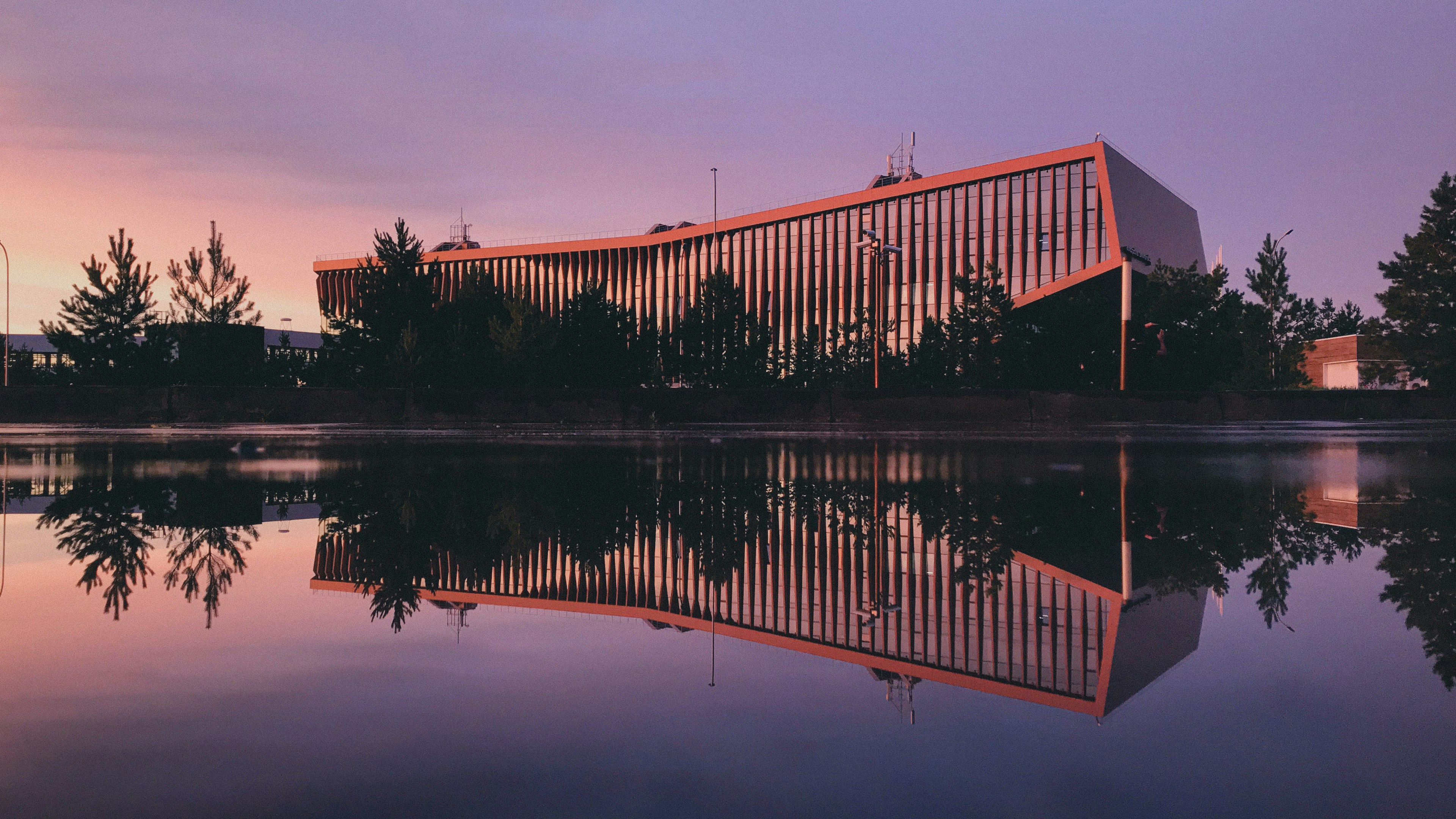 Modern architectural structure reflecting in calm water during twilight, surrounded by trees.