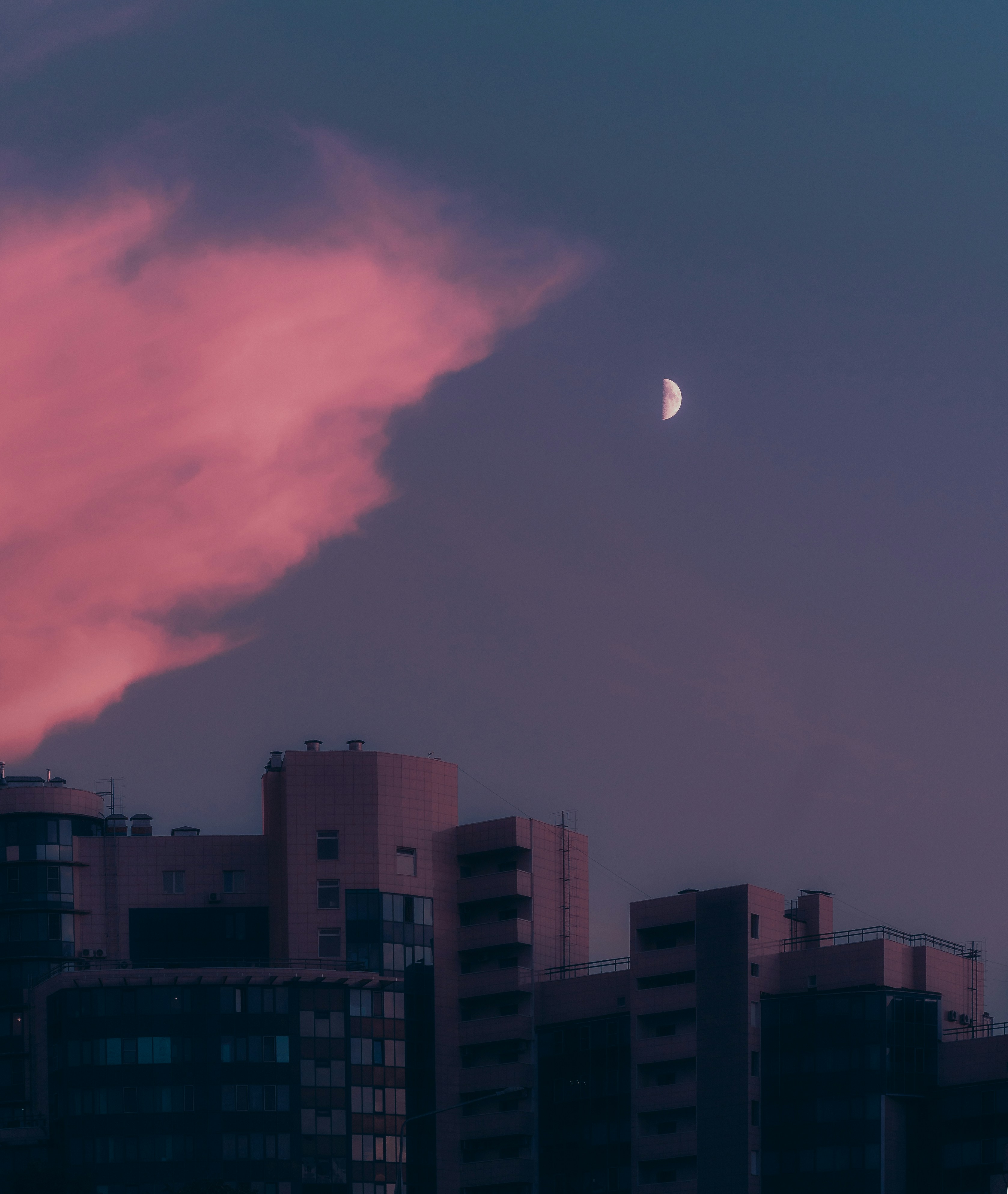 Soft pink clouds drift over modern architecture as a crescent moon hangs in the evening sky.