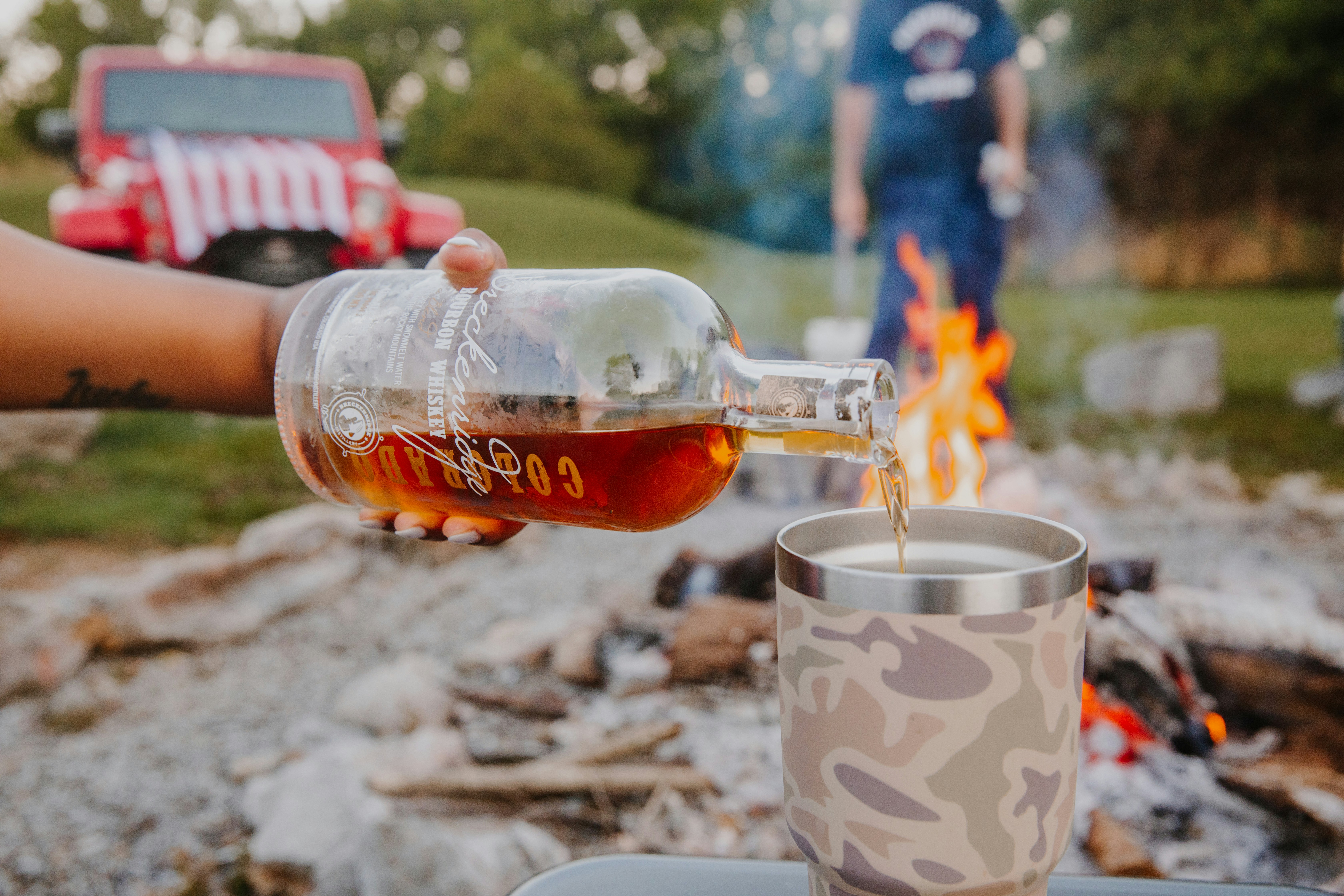 Bottle Pour Over a Campfire Toast: amber liquid streams into a camouflaged mug
