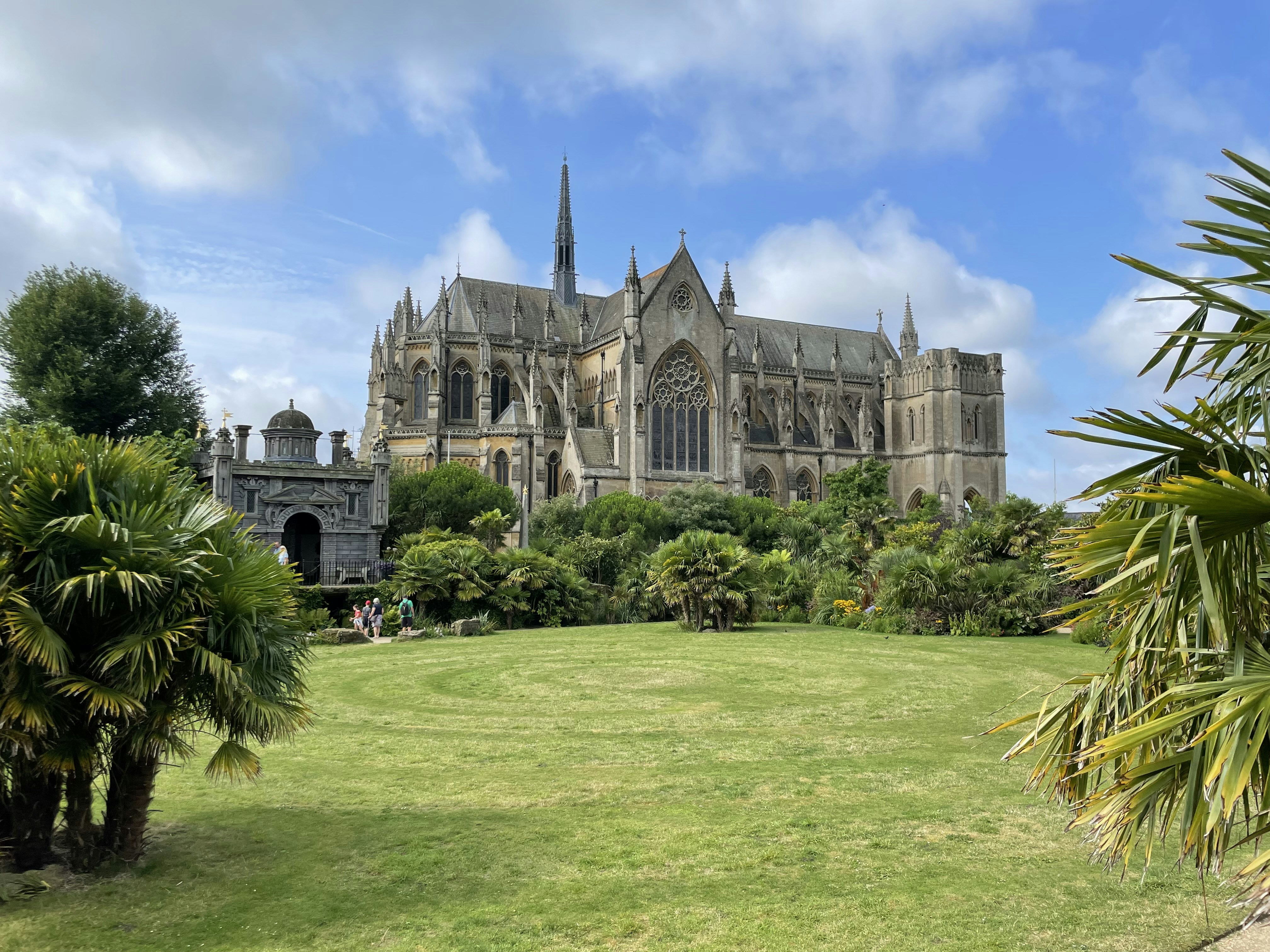 Gothic cathedral stands majestically behind vibrant palm trees in a tranquil garden setting. Visitors can be seen exploring the area.