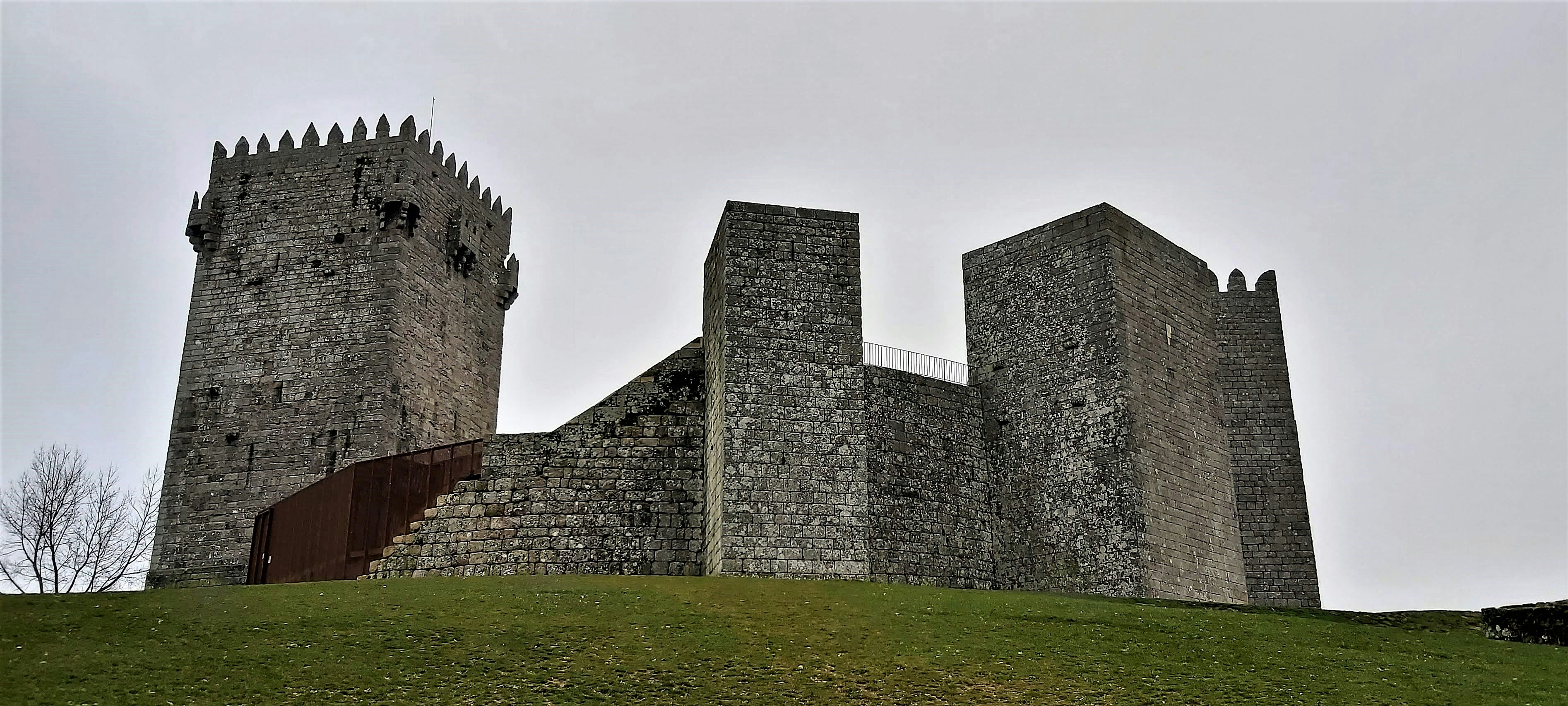Ruin of a medieval stone fortress rises on a grassy slope beneath a gray, overcast sky.